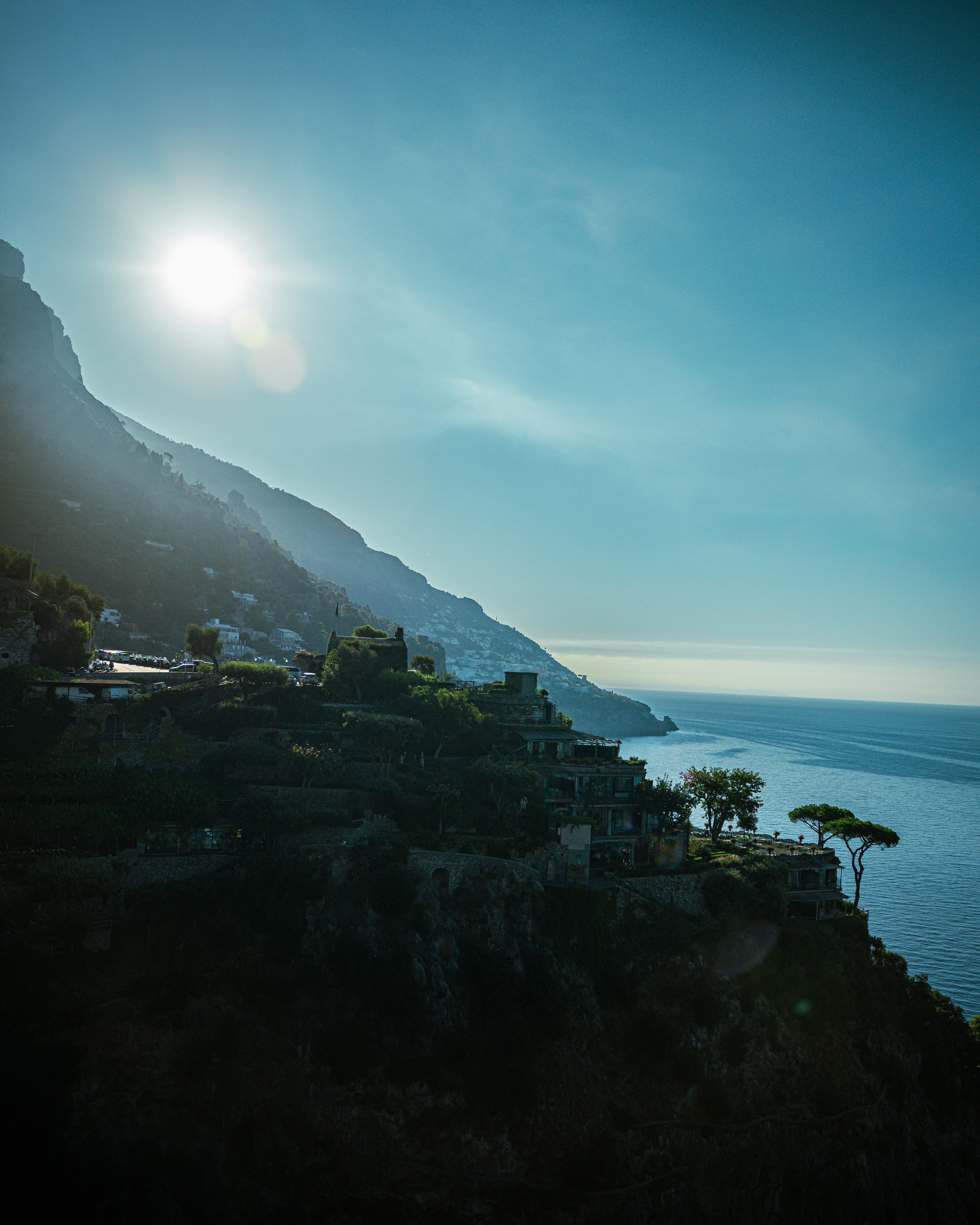 Coastal landscape showcasing terraced homes on a cliffside under a bright sun, with the serene sea stretching into the horizon.