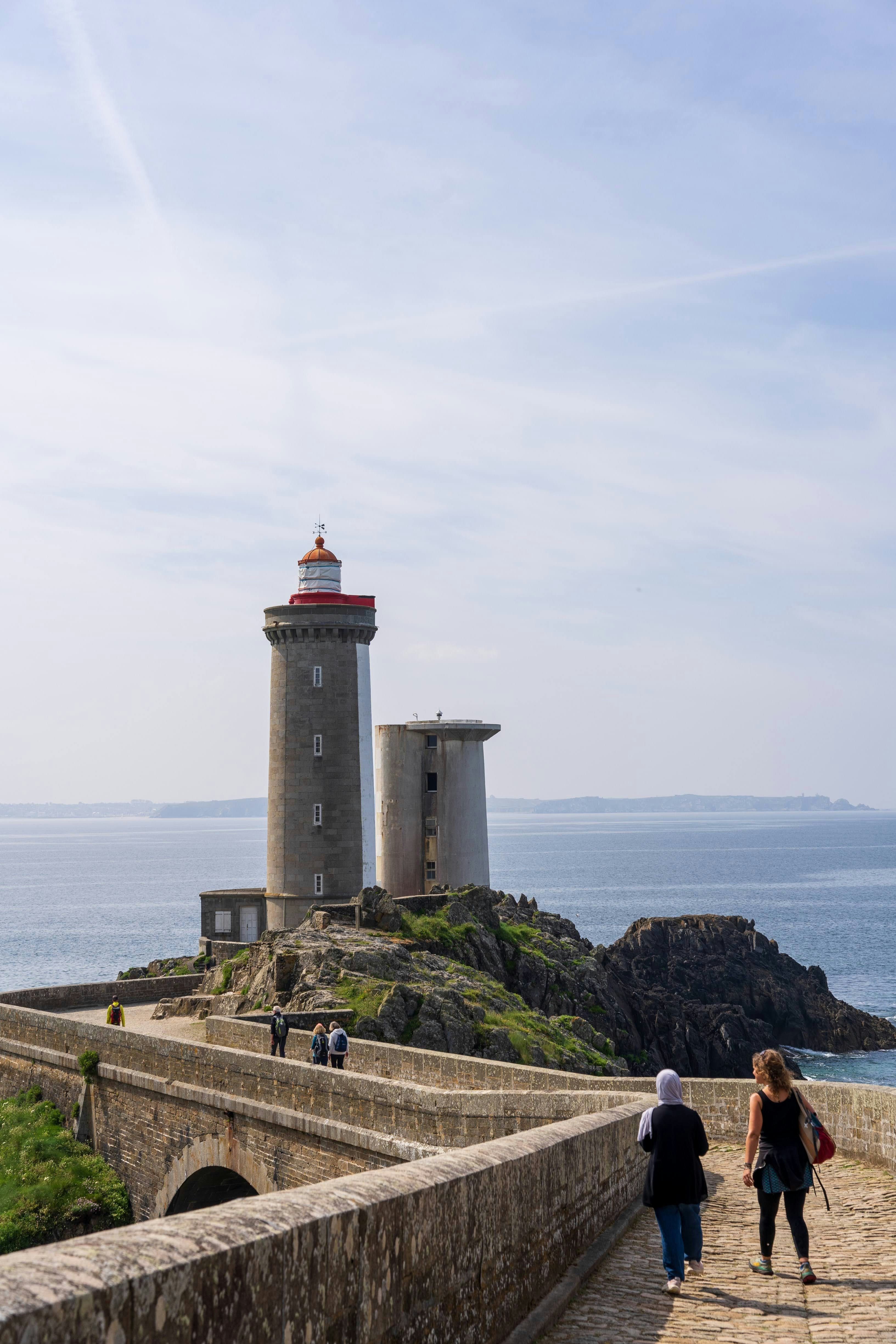 Two people walk towards a lighthouse on a rocky coast.