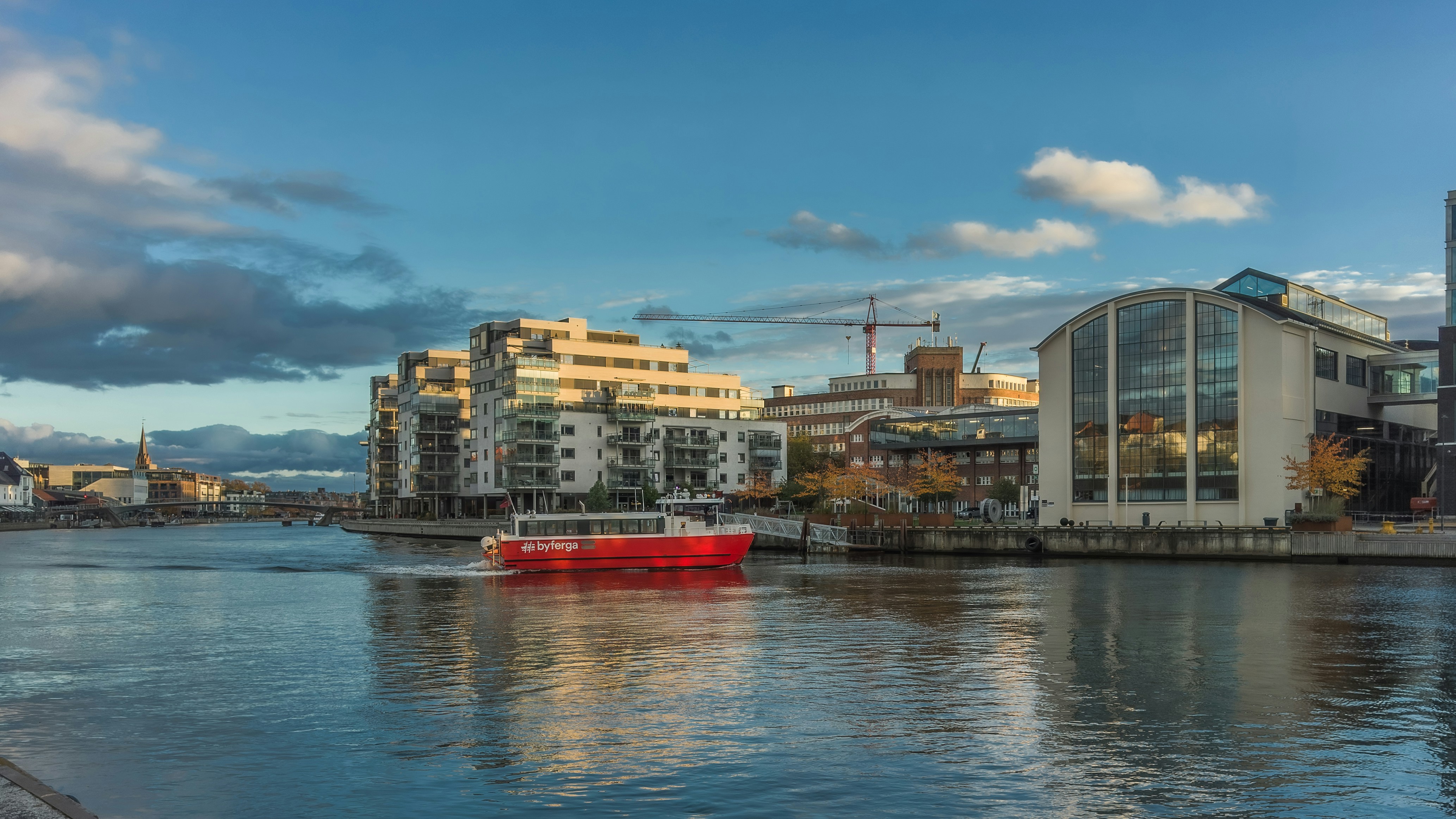 A vibrant red boat glides along the river, flanked by modern architecture and autumn foliage, capturing the essence of urban life. The scene is illuminated by a soft evening light.