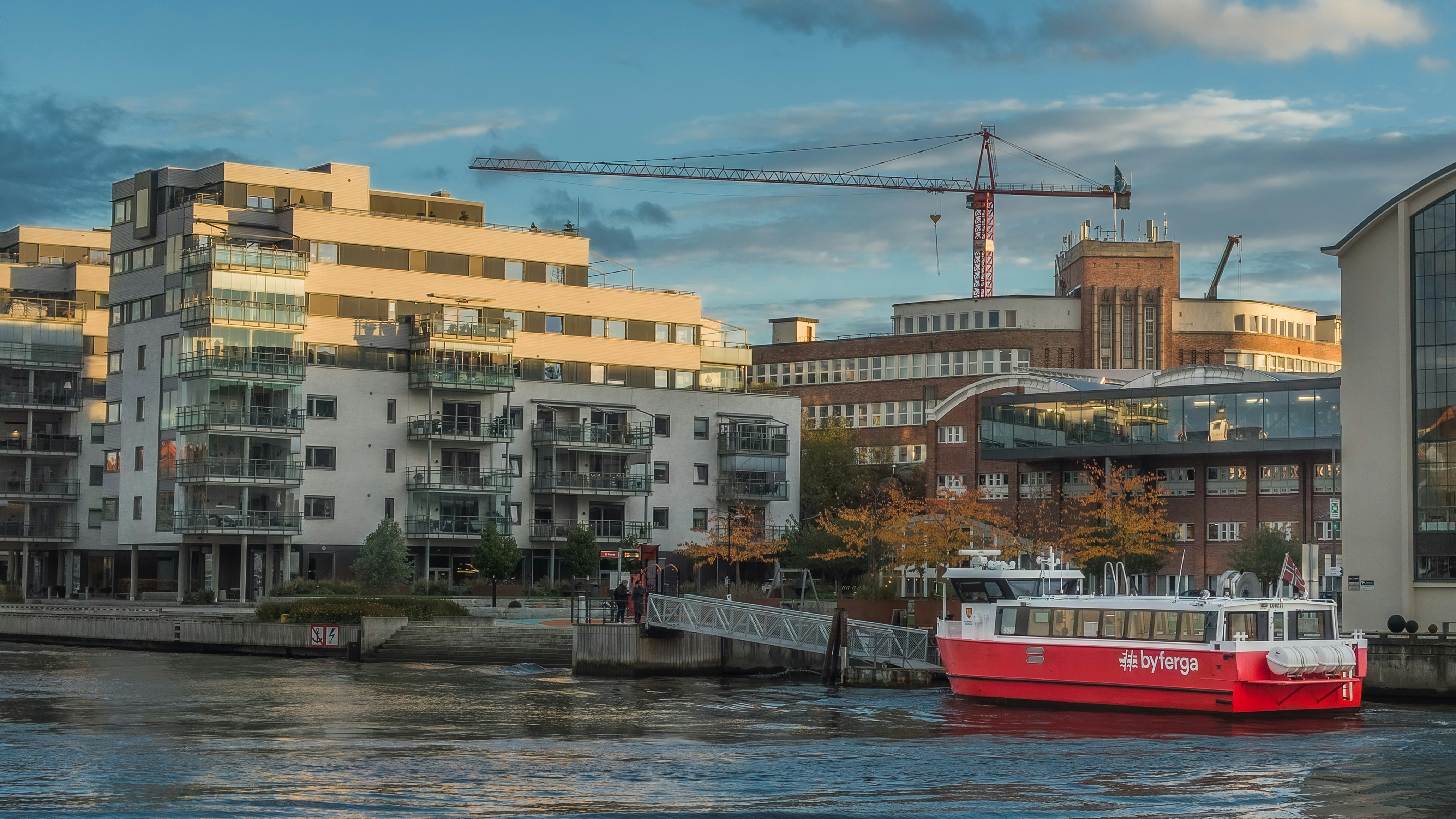 Bright red boat docked by modern waterfront buildings under a cloudy sky. The scene captures a blend of urban architecture and natural elements.