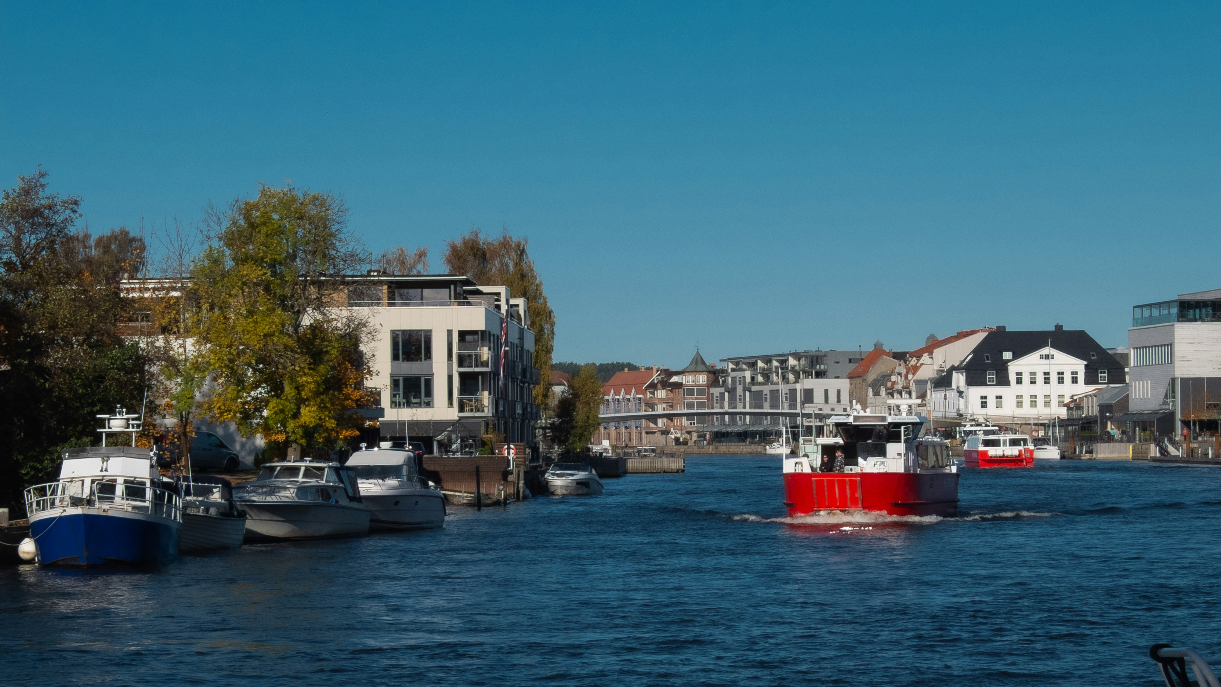 A vibrant red boat navigates through a bustling harbor lined with various vessels and autumn foliage, showcasing the lively atmosphere of riverside life.