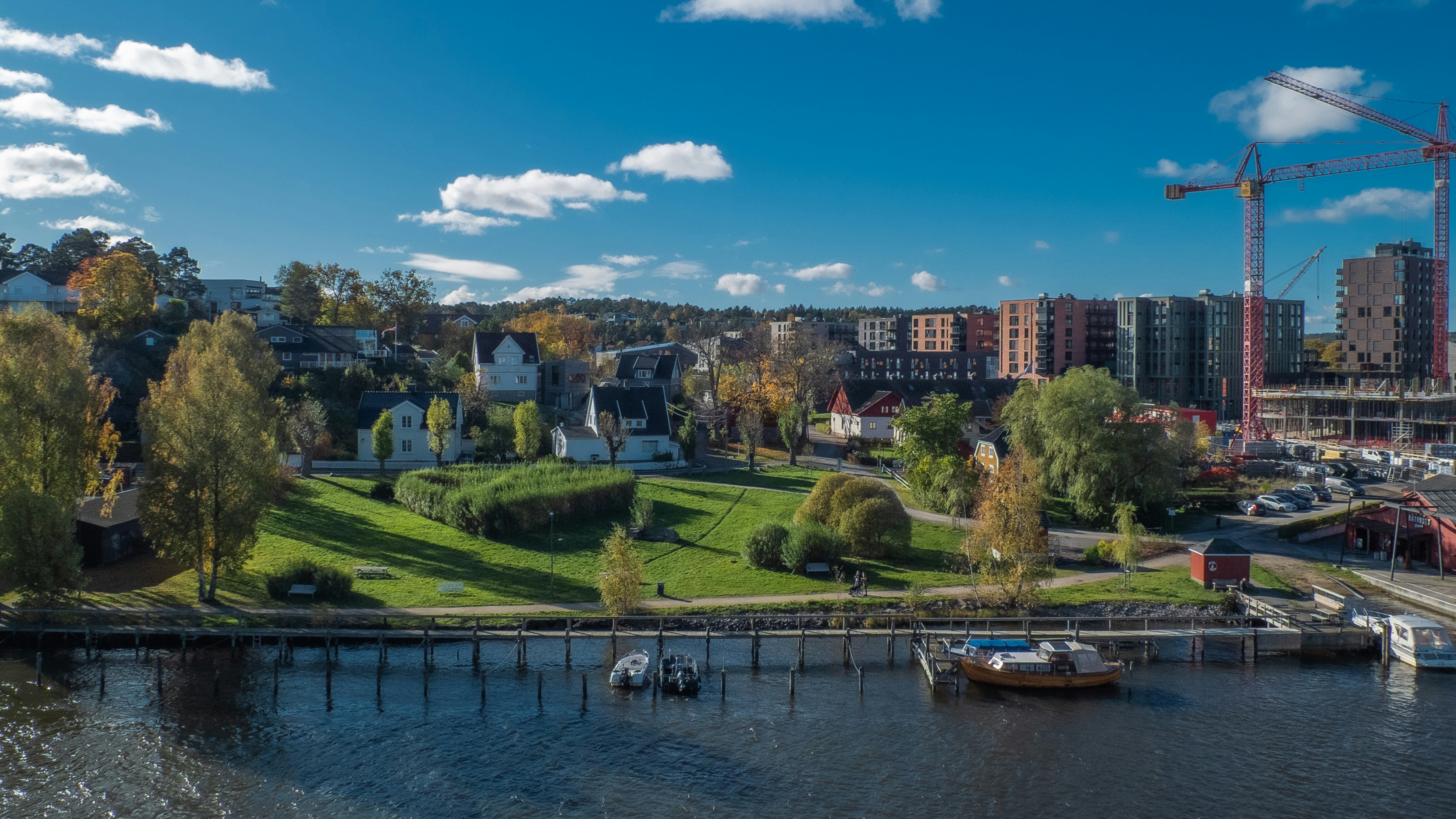 Colorful houses nestled among trees beside a serene harbor, with modern buildings and cranes in the background. Autumn foliage adds warmth to the scene.