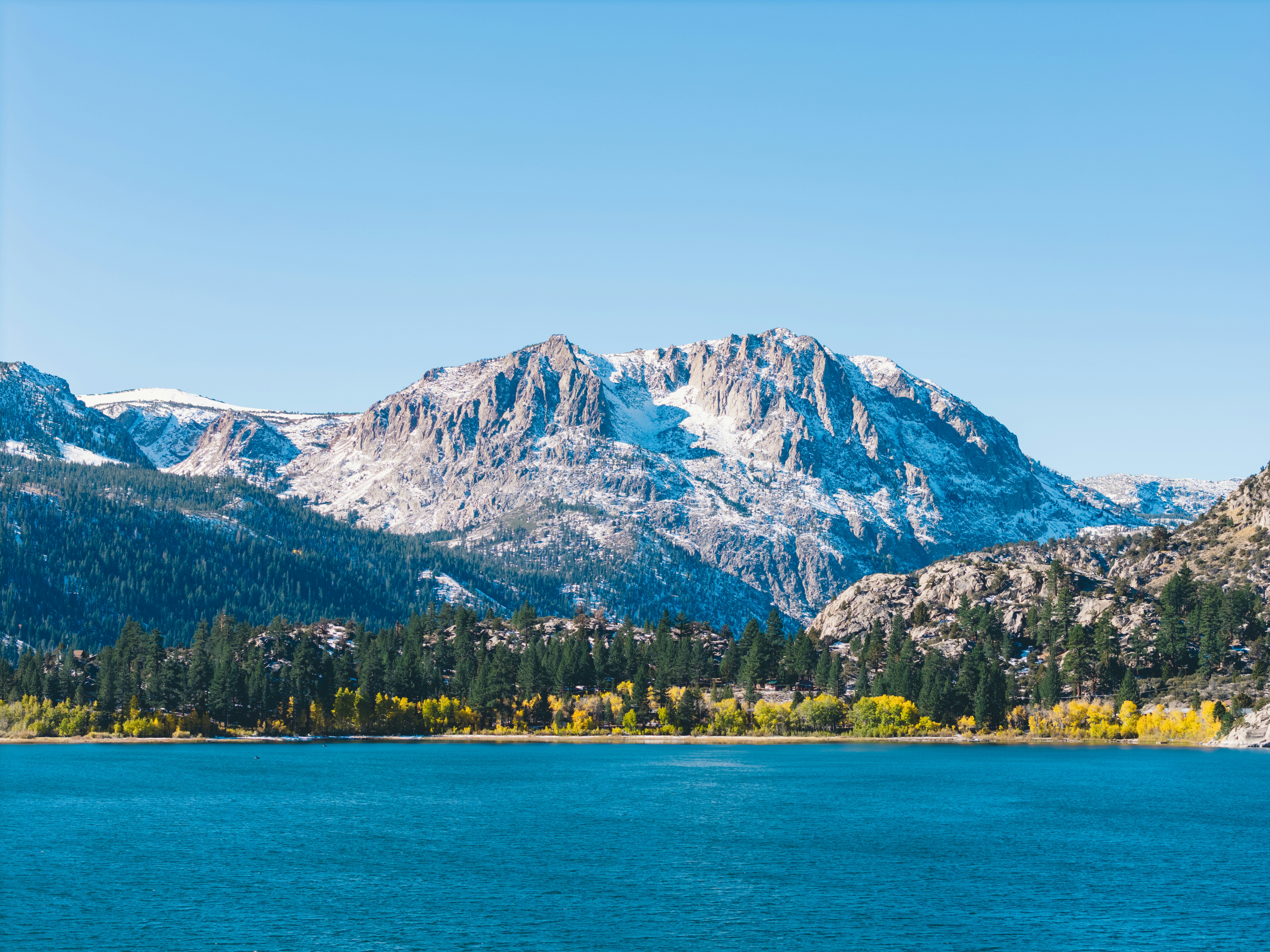 Snow-capped mountains rise majestically above a serene lake, framed by vibrant autumn foliage. The clear blue sky enhances the tranquil scene.