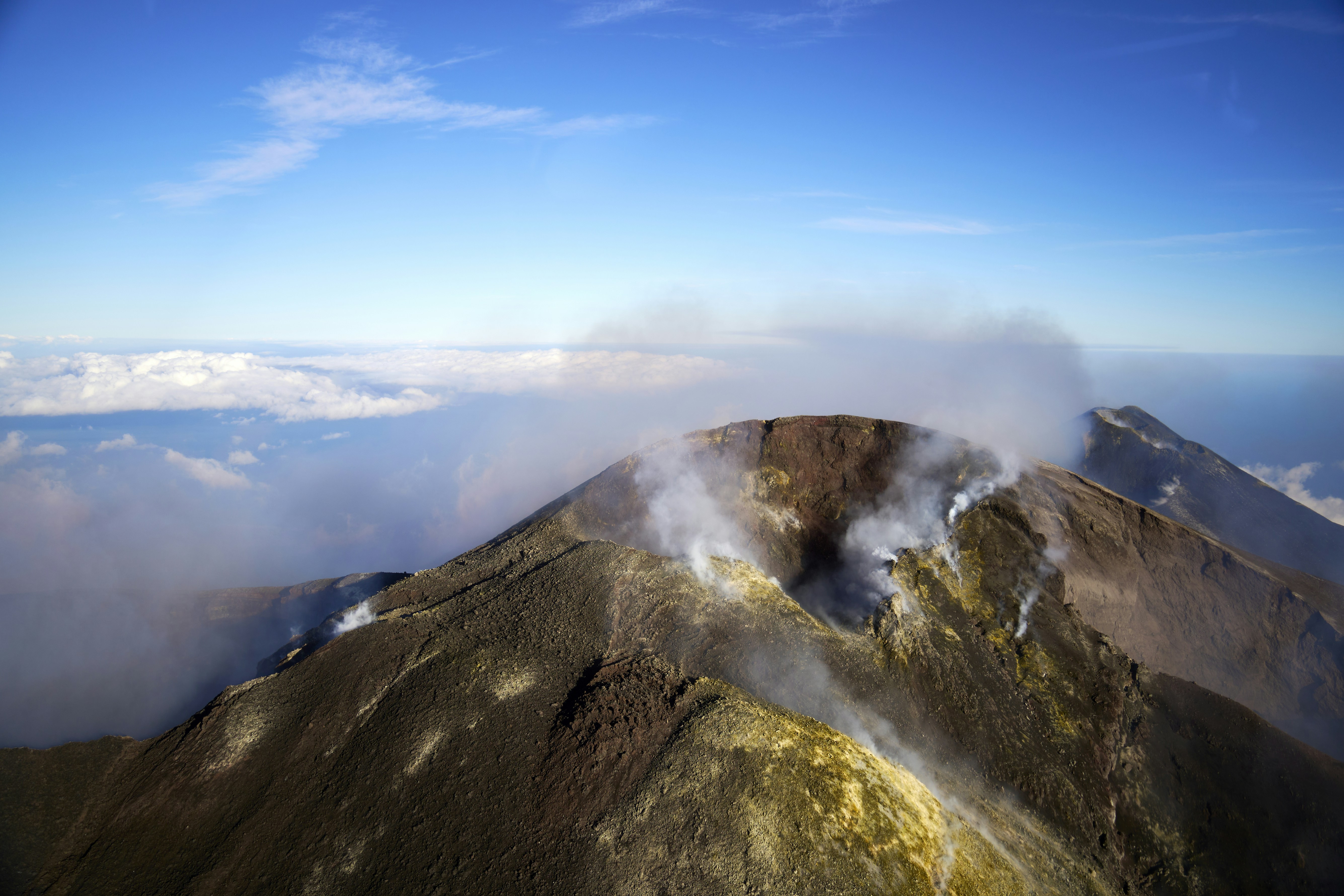 Aerial view of a volcanic mountain with smoke gently rising from its peak, set against a backdrop of blue sky and distant clouds.
