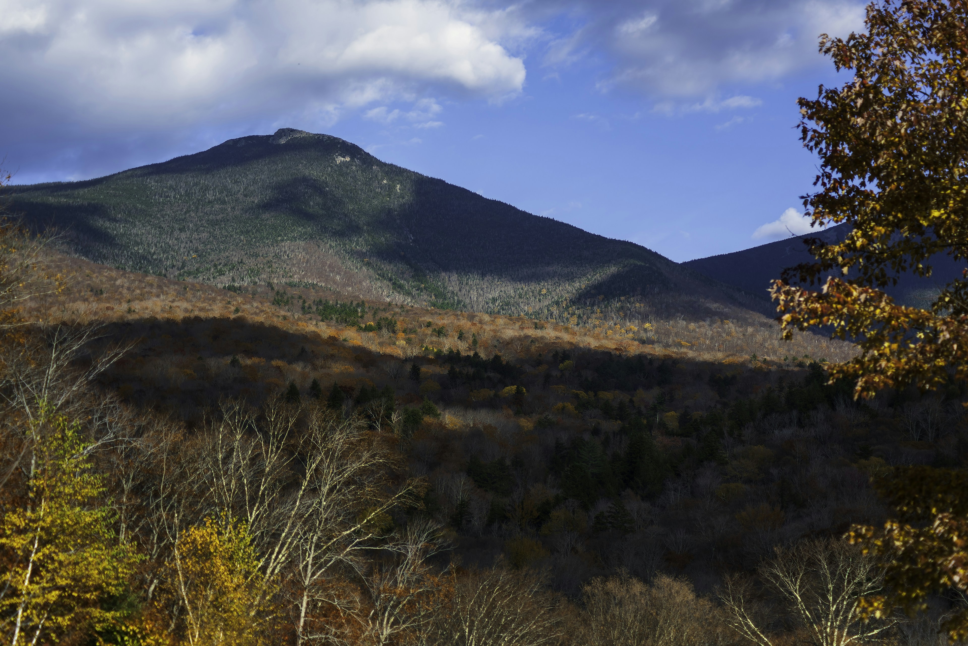 Vibrant autumn foliage blankets the foothills beneath a majestic mountain, illuminated by soft sunlight breaking through scattered clouds.