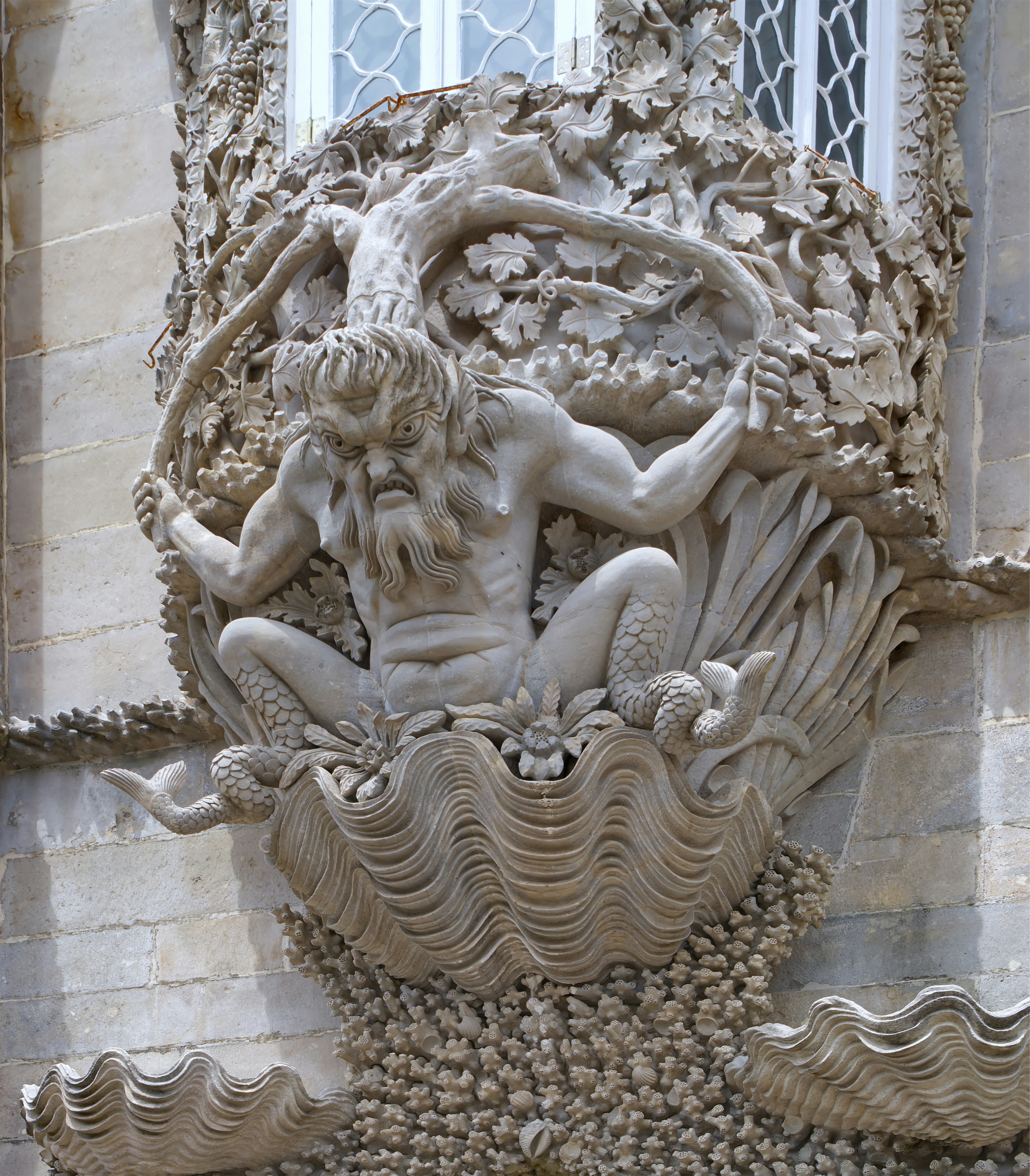 Au sommet d’une des portes majestueuses du Palais de Pena, ce Triton, gardien des mers et messager des profondeurs, se dresse dans toute sa puissance mythique. Son visage, marqué par des traits expressifs et une barbe sauvage, incarne la force brute et la sagesse ancienne des océans. Les yeux écarquillés, les sourcils froncés et la bouche entrouverte semblent lancer un appel aux éléments, comme s’il commandait aux vagues et aux vents. | Stone sculpture of a merman in a shell