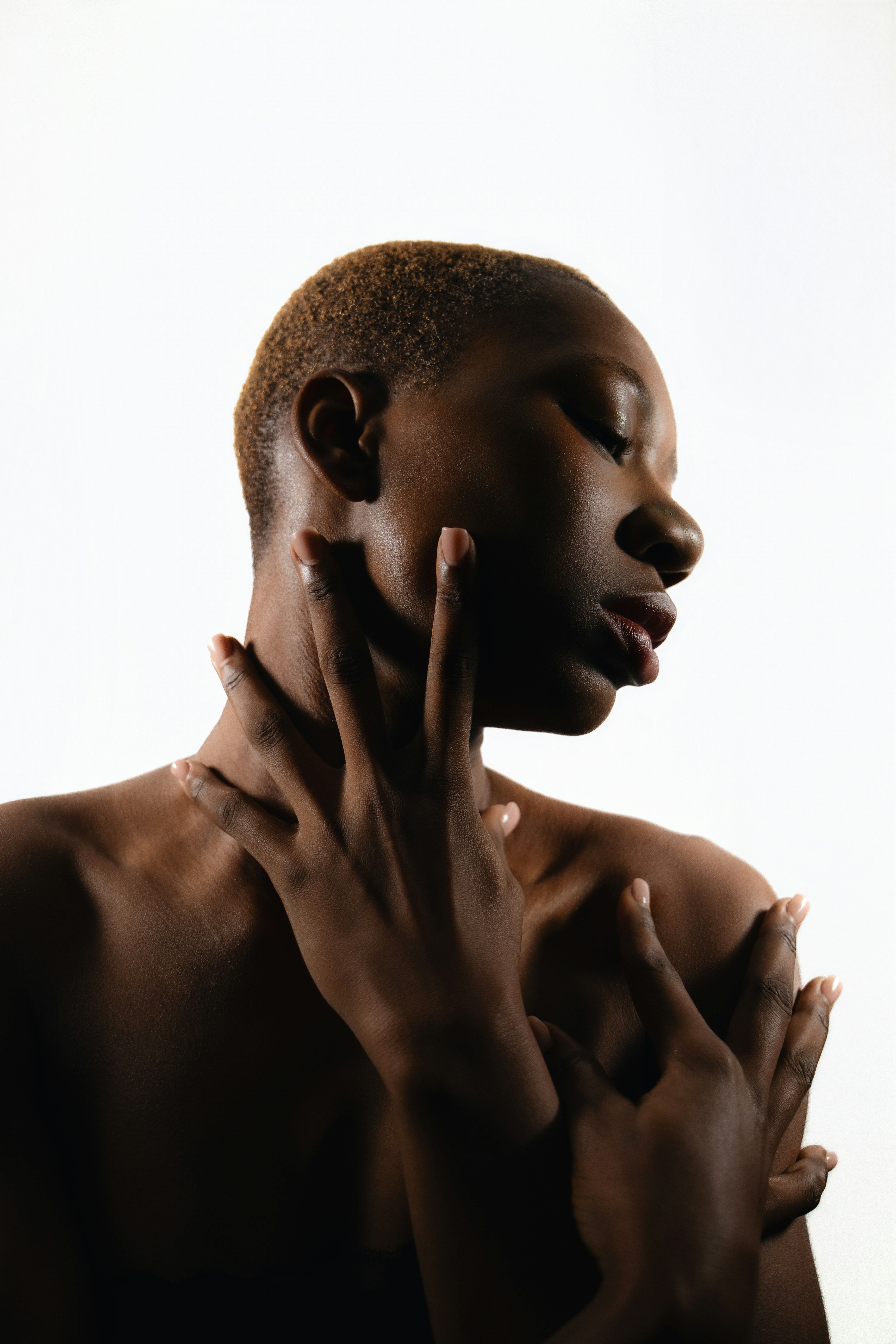 A profile of a person with short hair, delicately holding their neck with both hands against a stark white background.