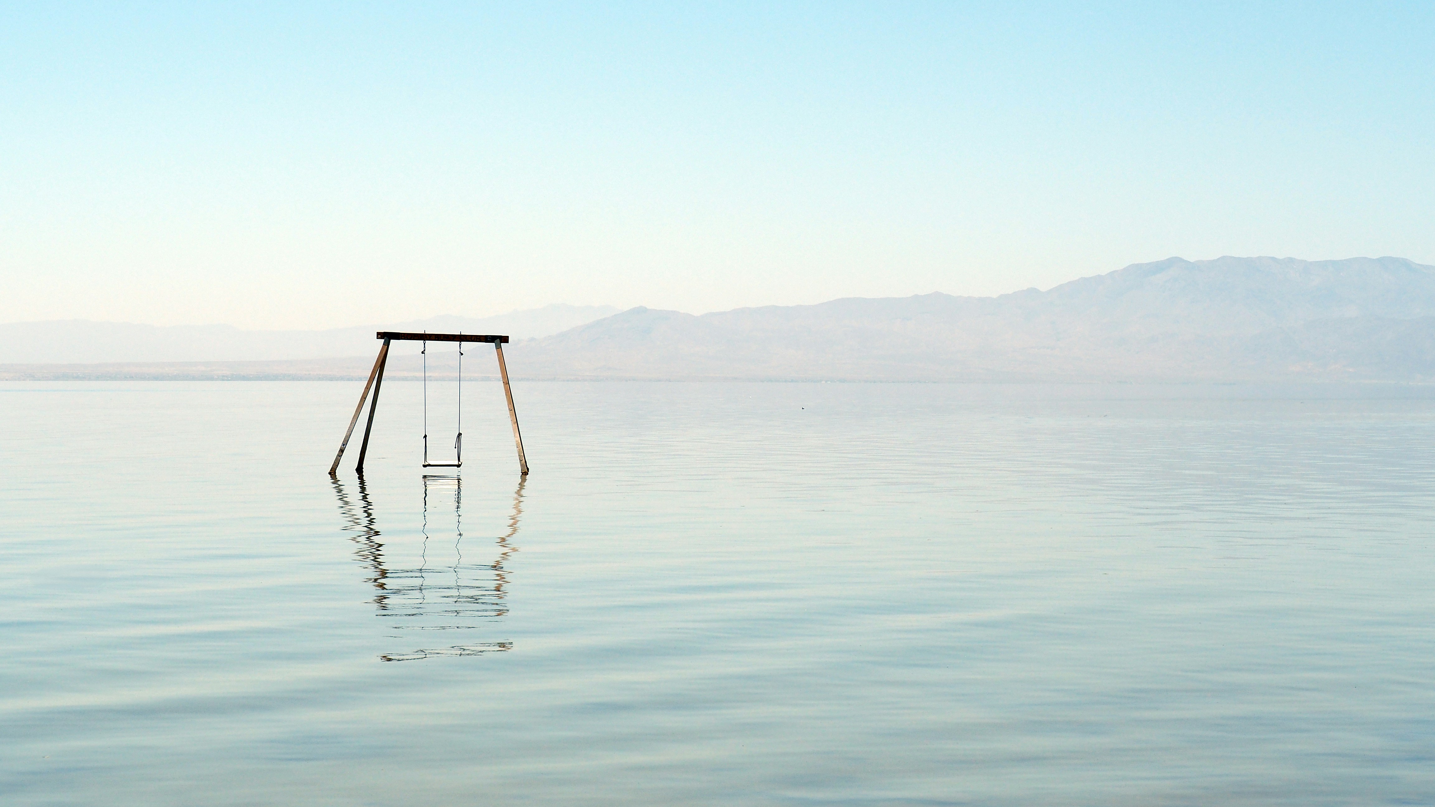 OLYMPUS DIGITAL CAMERA | A swing set stands in calm water with mountains afar.