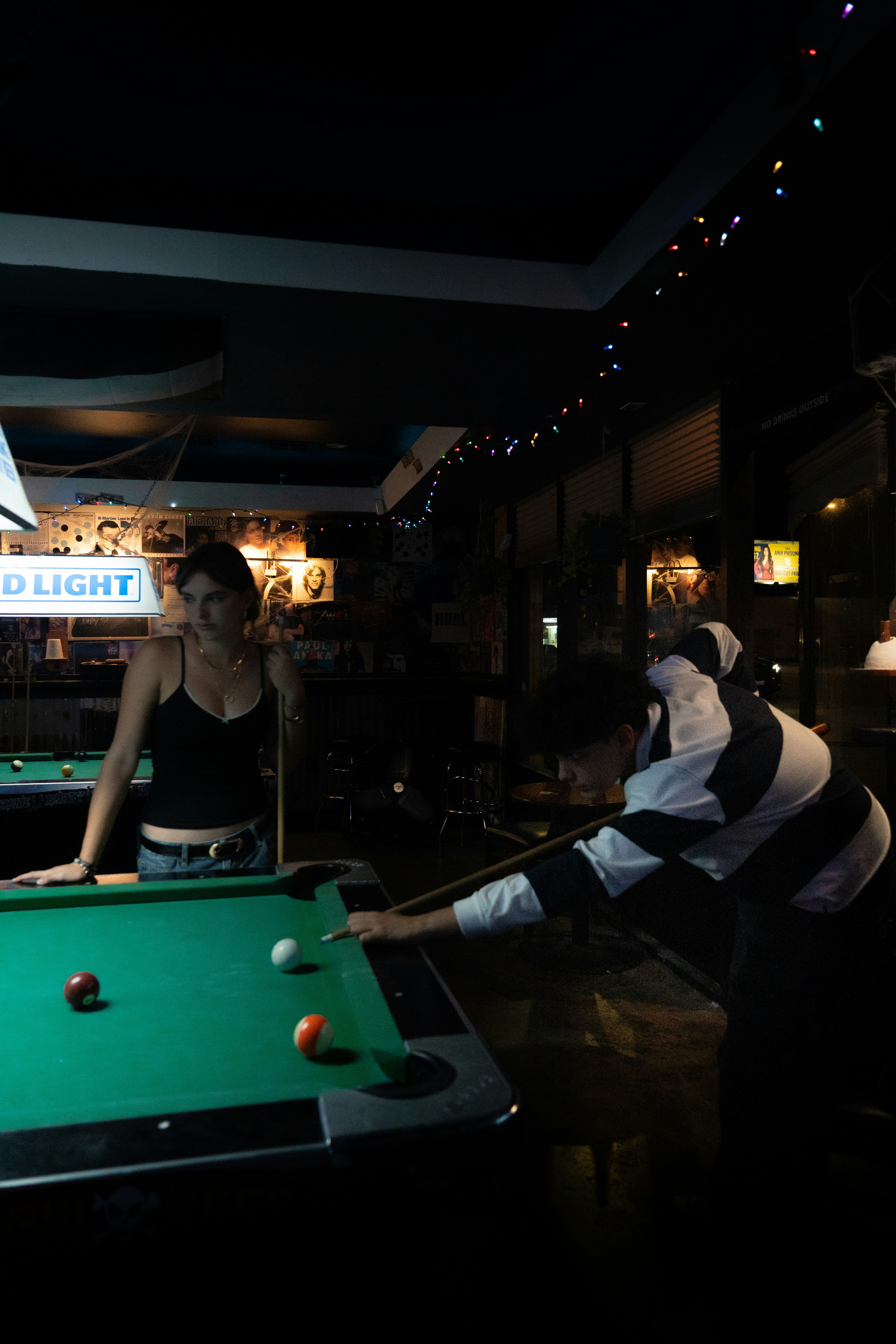 Two individuals engaged in a game of pool under dim lighting, with a 'Bud Light' sign illuminating the scene.