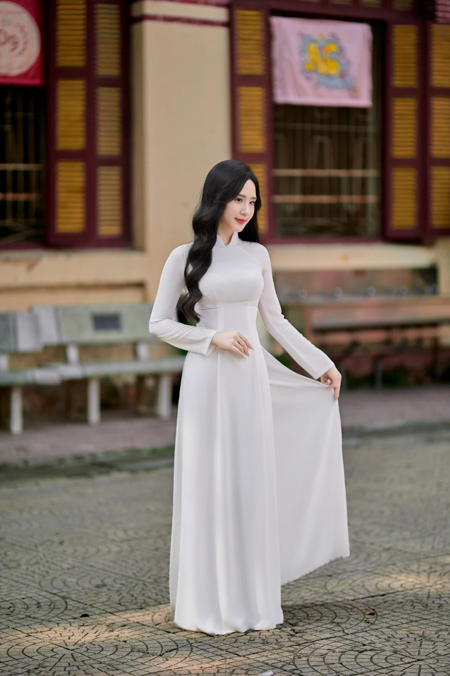Woman in a flowing white áo dài standing outdoors, holding the fabric of her dress with traditional buildings softly blurred behind her.