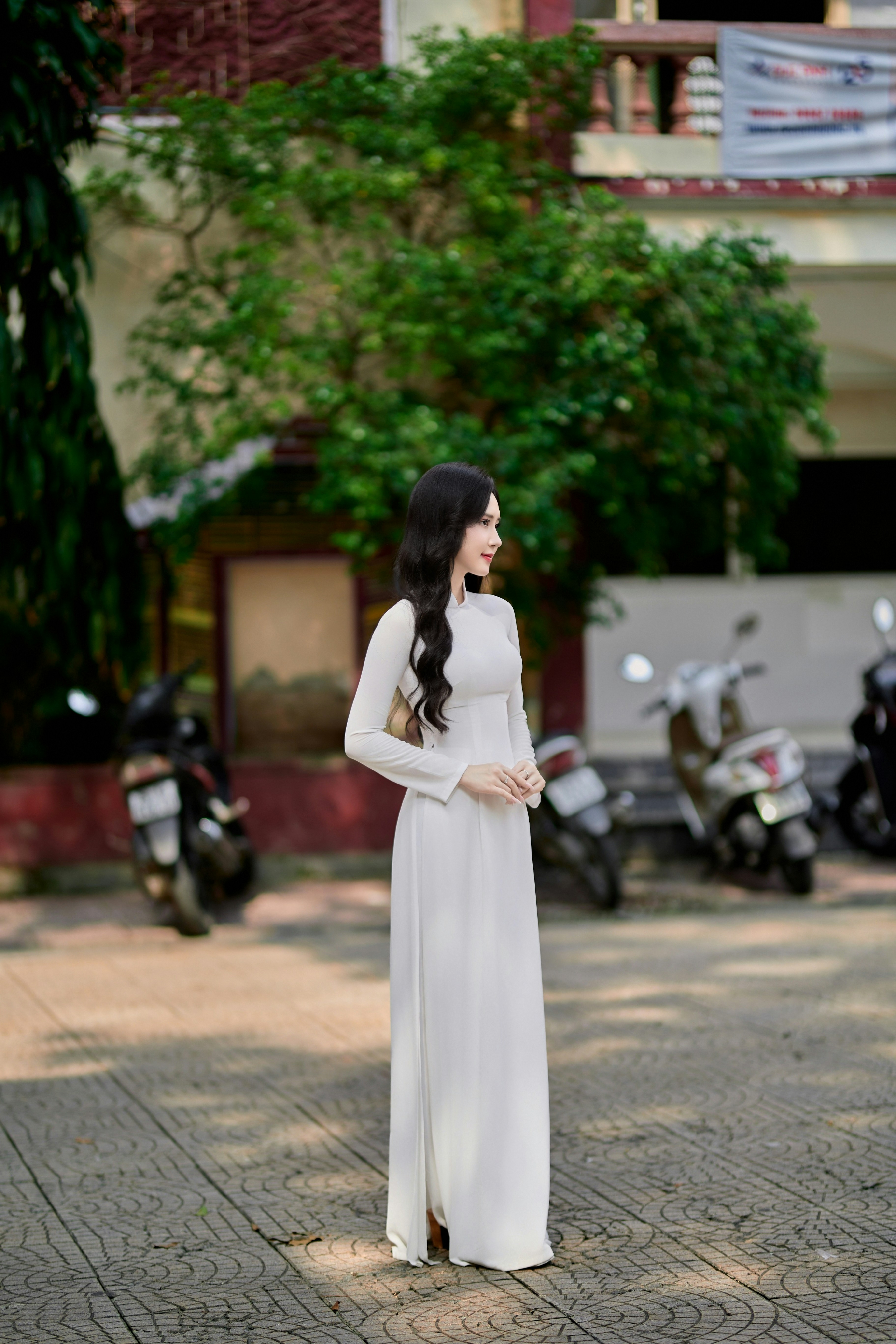 A woman in a flowing white dress stands gracefully amidst a backdrop of greenery and parked motorcycles. Her serene expression complements the tranquil setting.