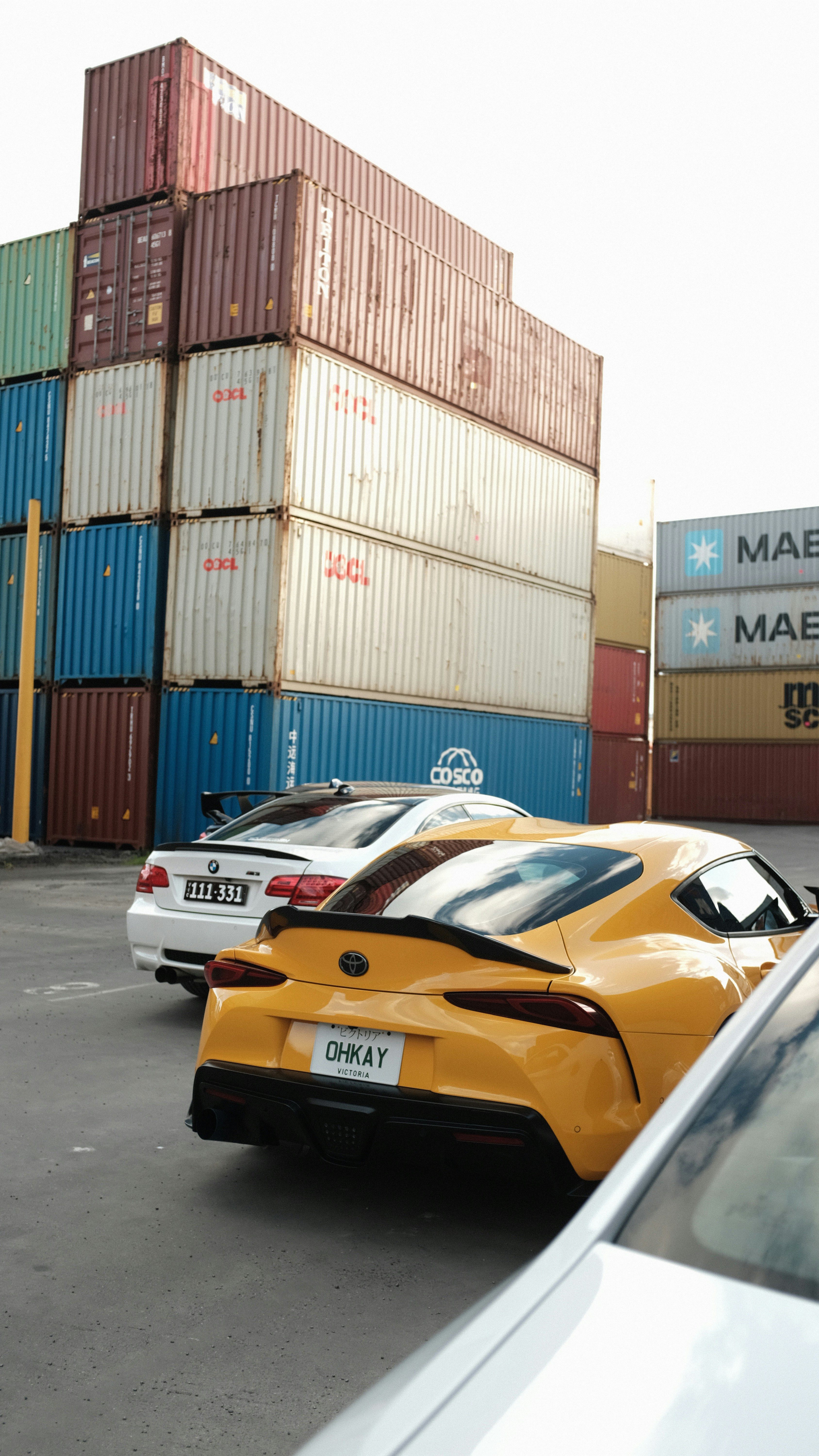 Two sports cars parked near stacked shipping containers.