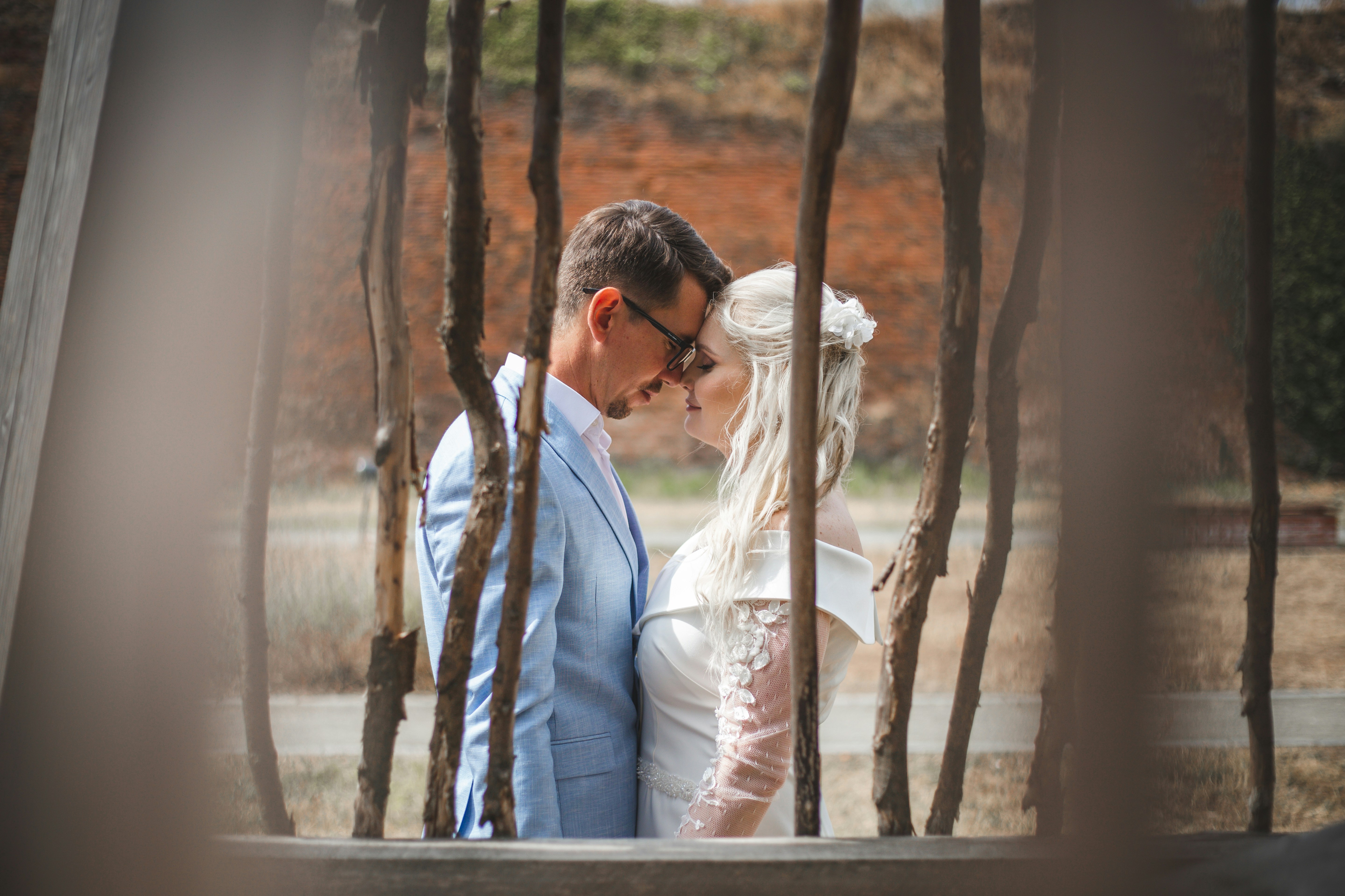 Man and his fiance close together, head to head, wood elements in front of them. | Couple embracing behind wooden bars