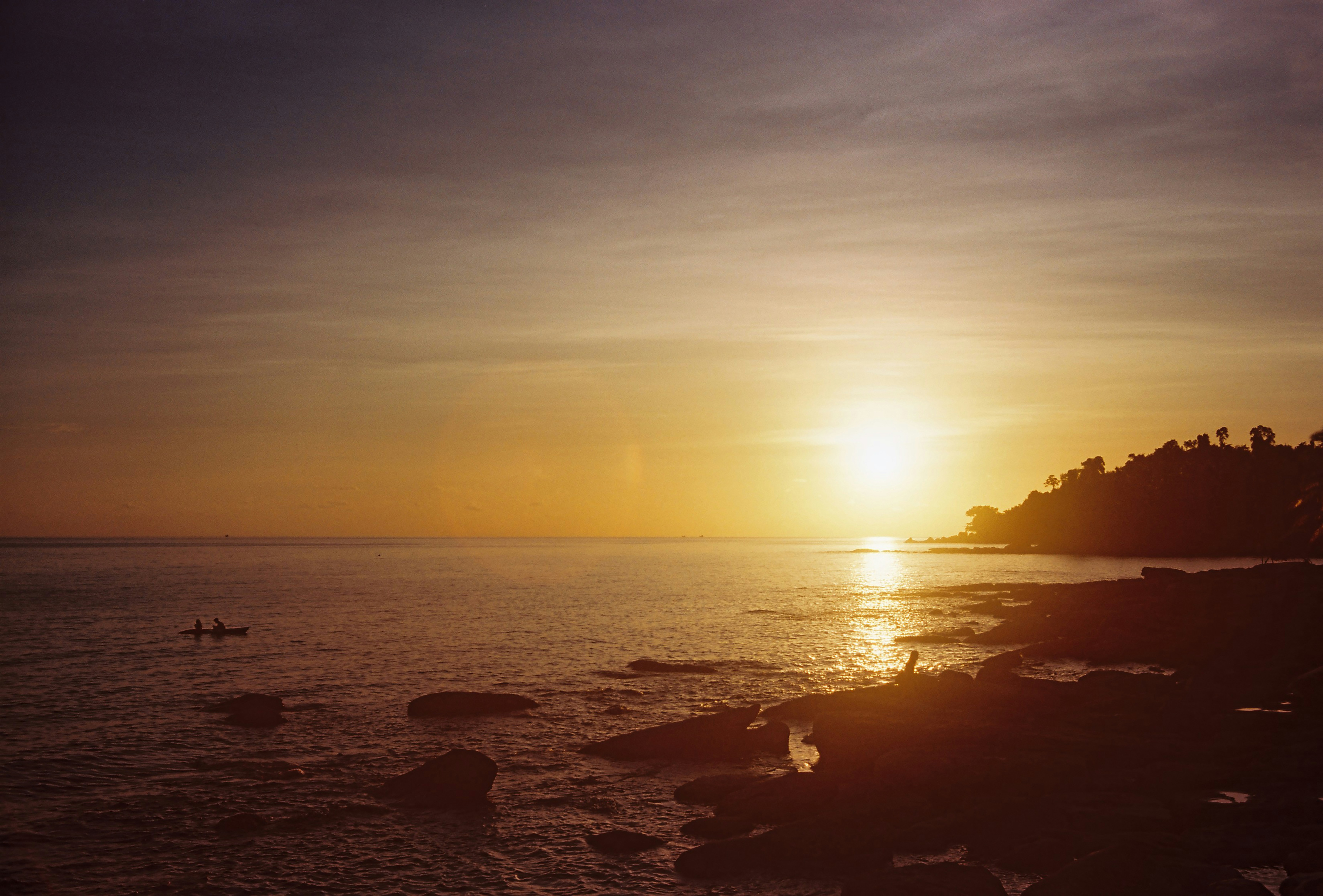 Kayakers glide across a tranquil sea as the sun sets behind a coastal silhouette, casting warm hues across the water.