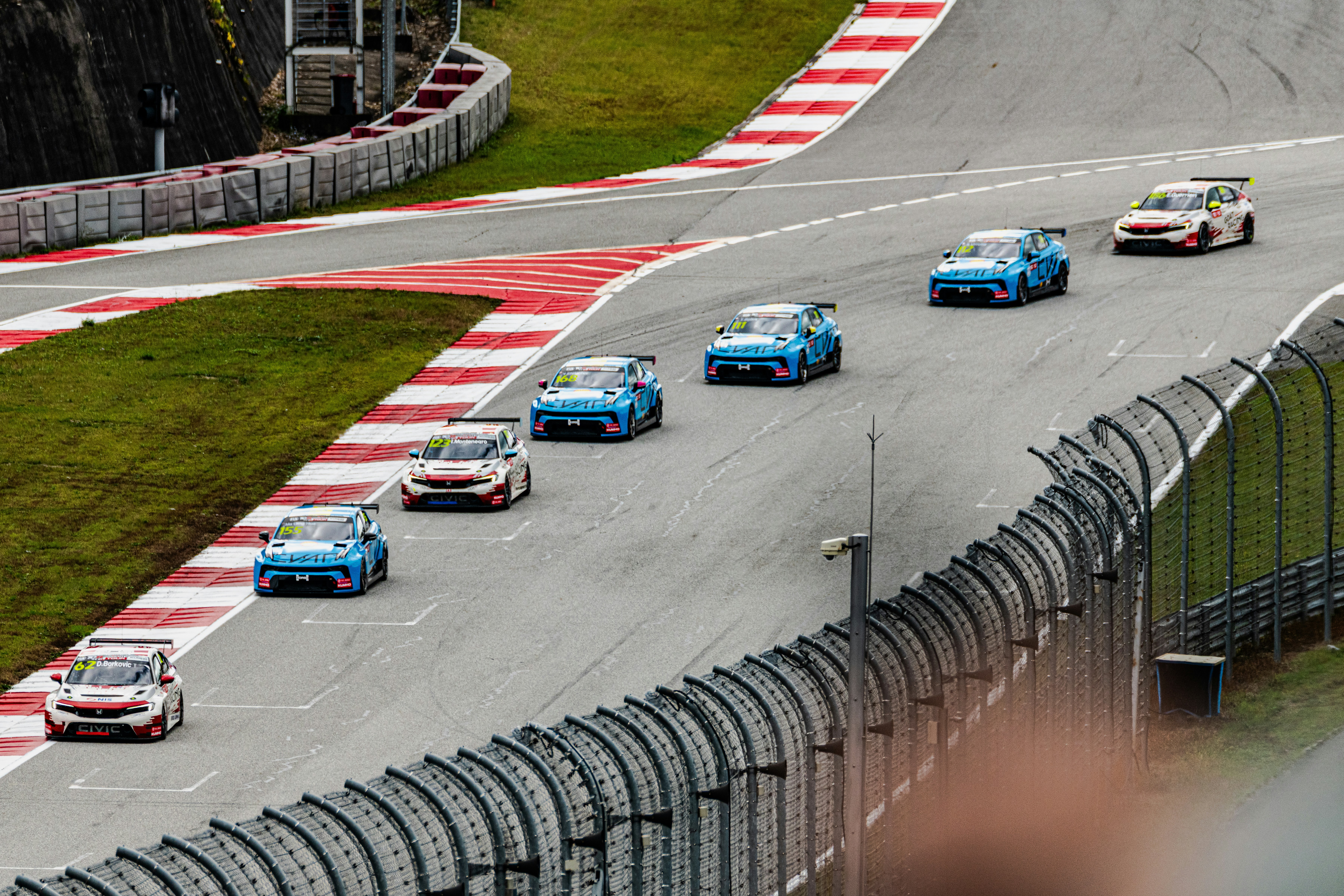 Six race cars navigate a sharp turn on a racing circuit, showcasing the thrill of competitive motorsport. The vibrant colors of the cars contrast with the track's red and white markings.
