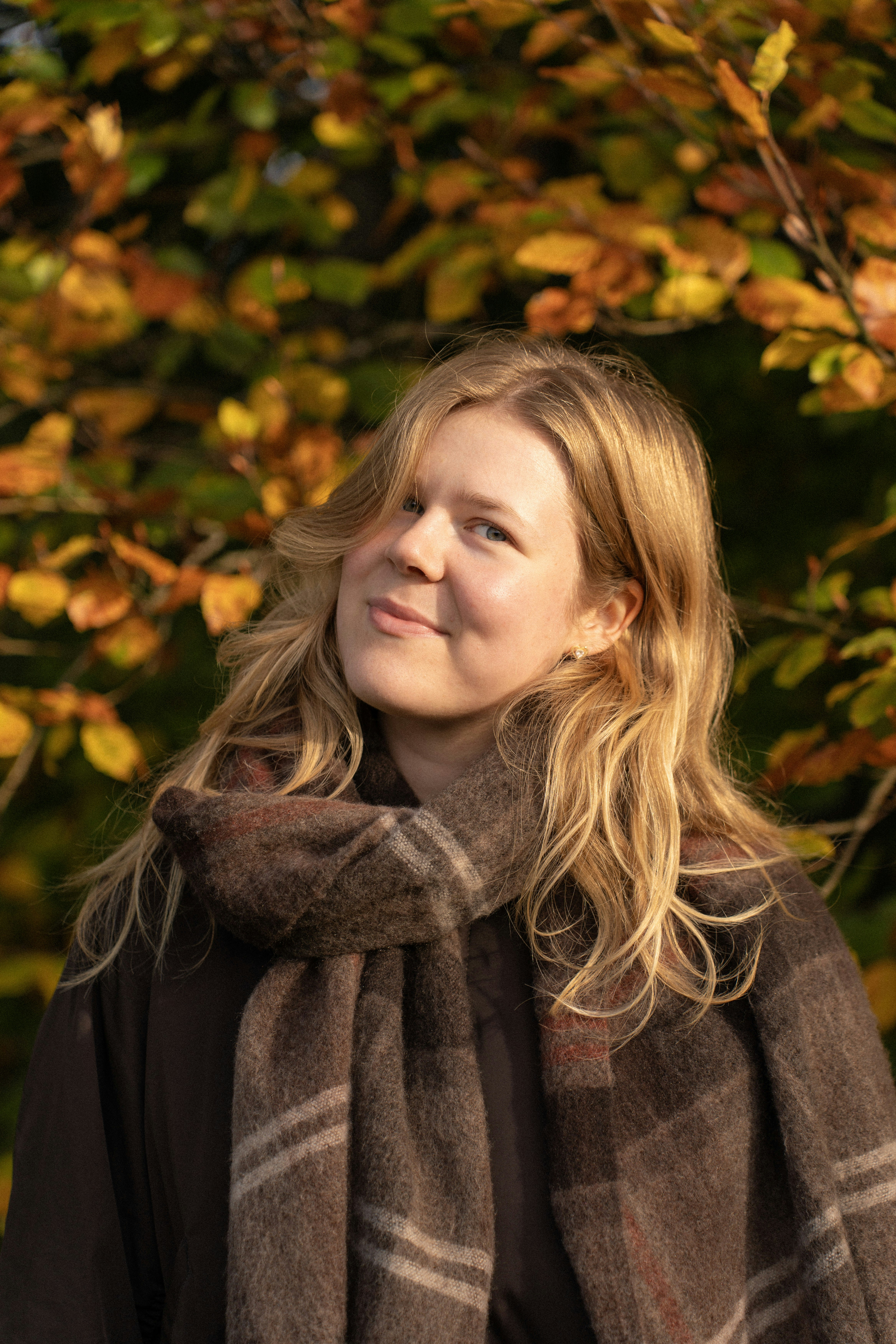 Young woman with flowing hair and a cozy scarf smiles softly against a backdrop of vibrant autumn foliage.