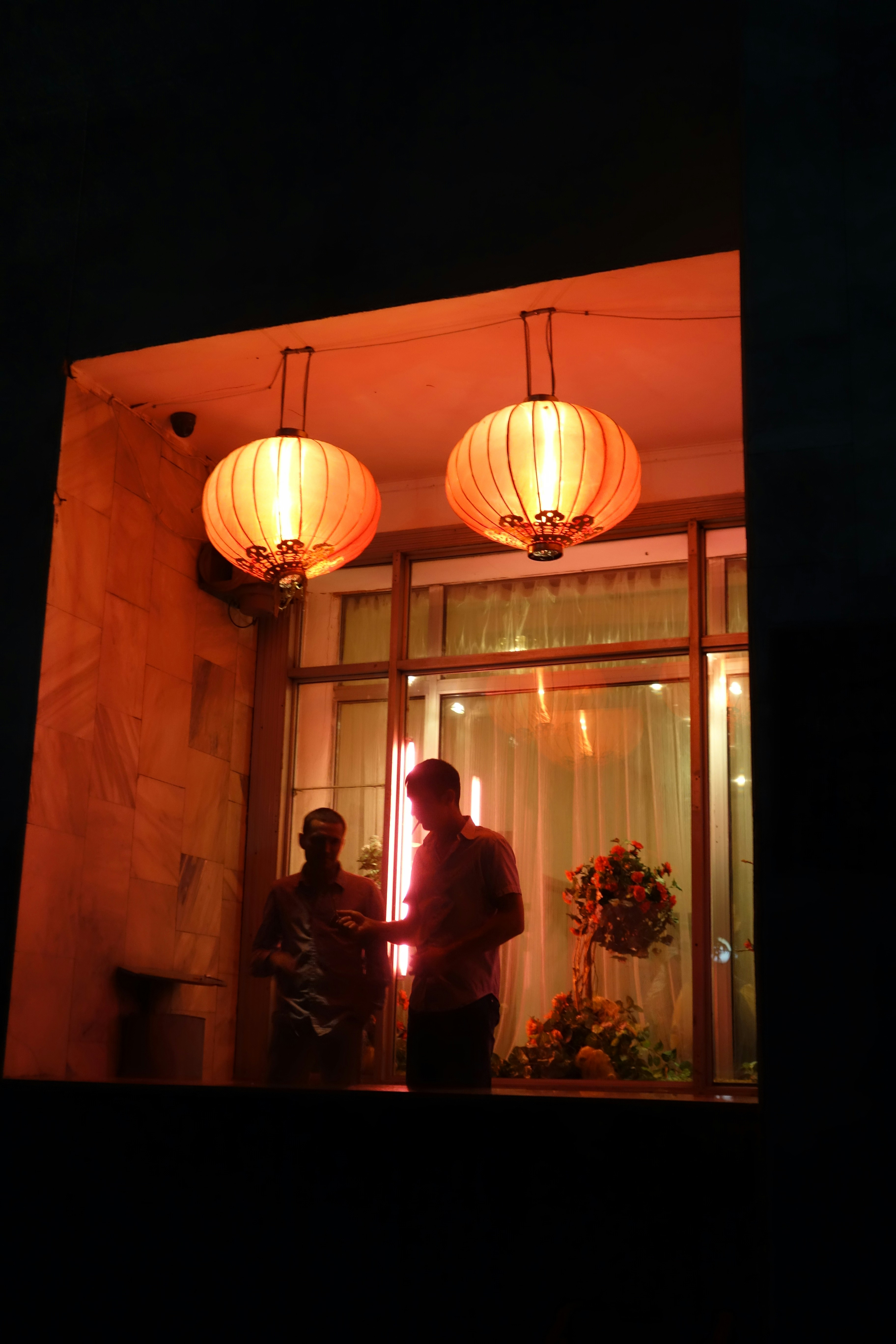 Two men stand near glowing lanterns at night.