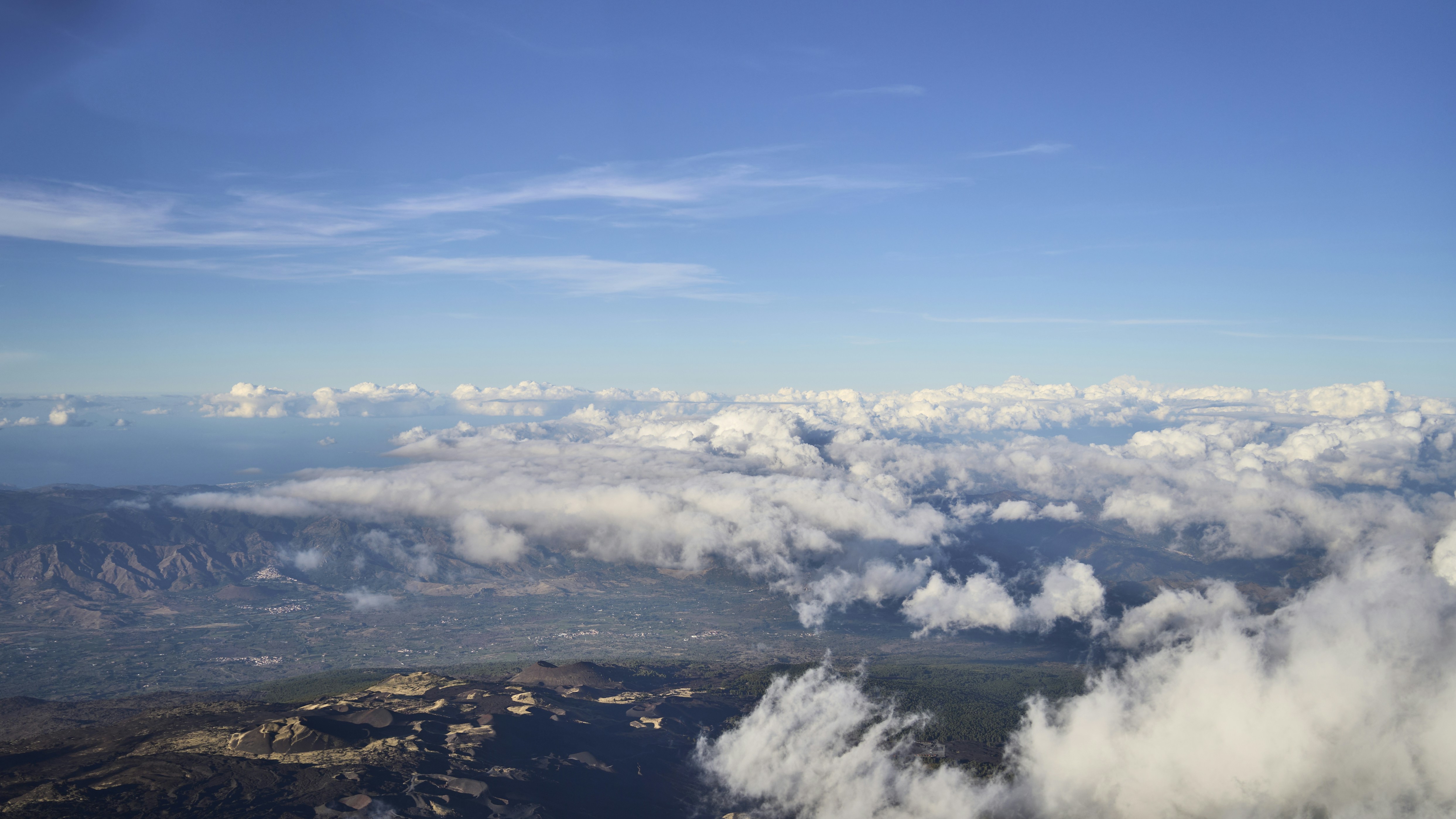 Wenn man schon mal am Ätna ist und hat die Chance auf einen Hubschrauber Flug über den Vulkan sollte man das machen, man lebt ja nur einmal. Wichtig nicht nur fotografieren sondern auch den Flug genießen Blick vom Vulkan Richtung Küste. | Clouds float over a mountainous landscape under blue sky.