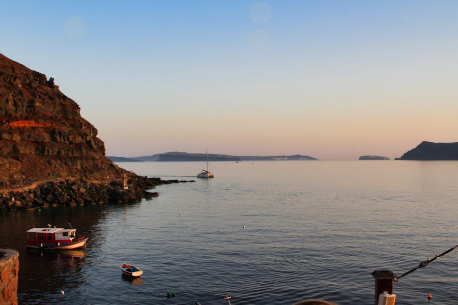 Small sailboat moored near a rocky cliff on calm water with distant islands under a pastel dusk sky.