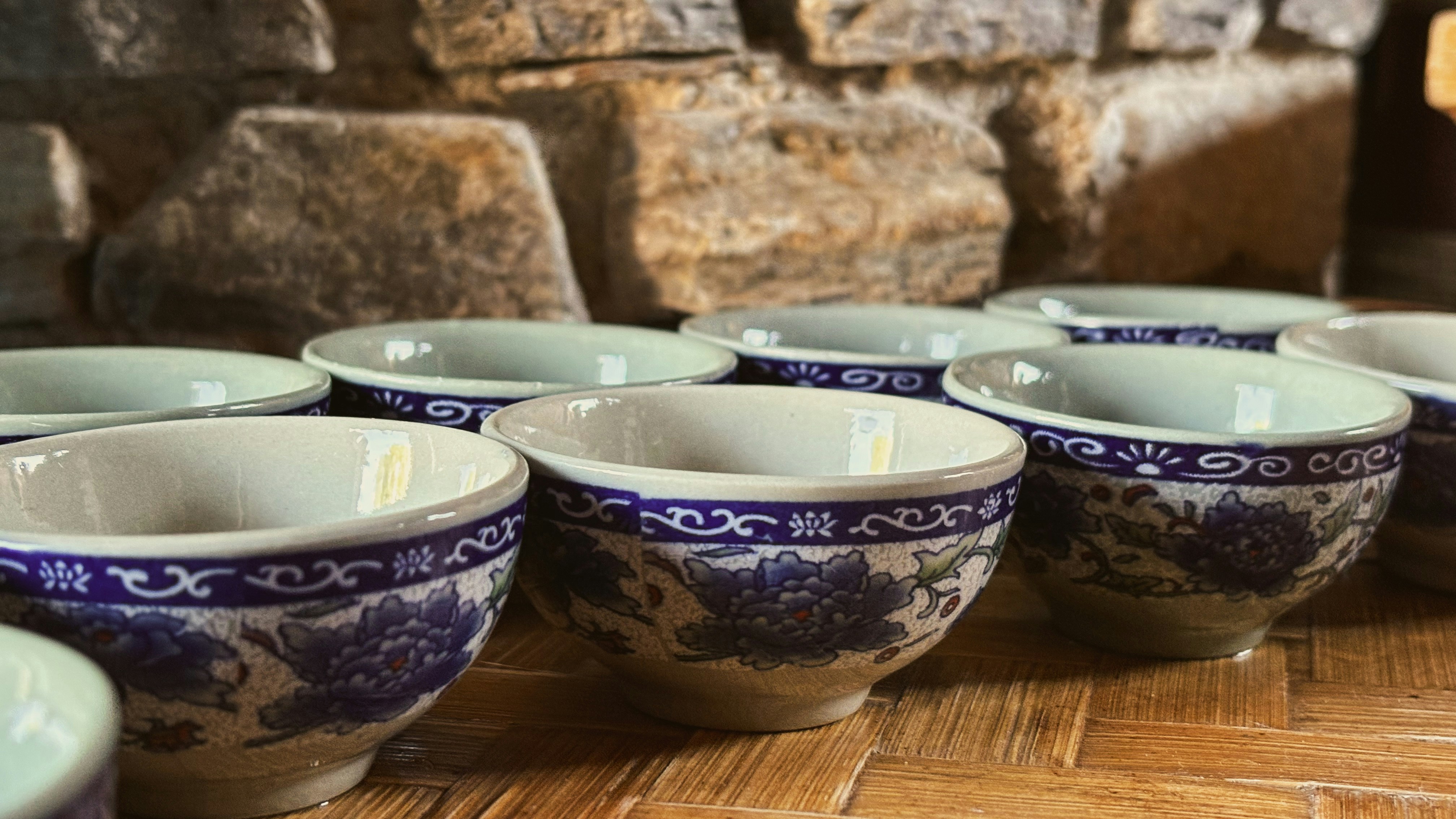 A row of hand-painted ceramic bowls with delicate floral designs rests on a wooden surface against a stone wall, reflecting the charm of Bhutanese craftsmanship and tradition.