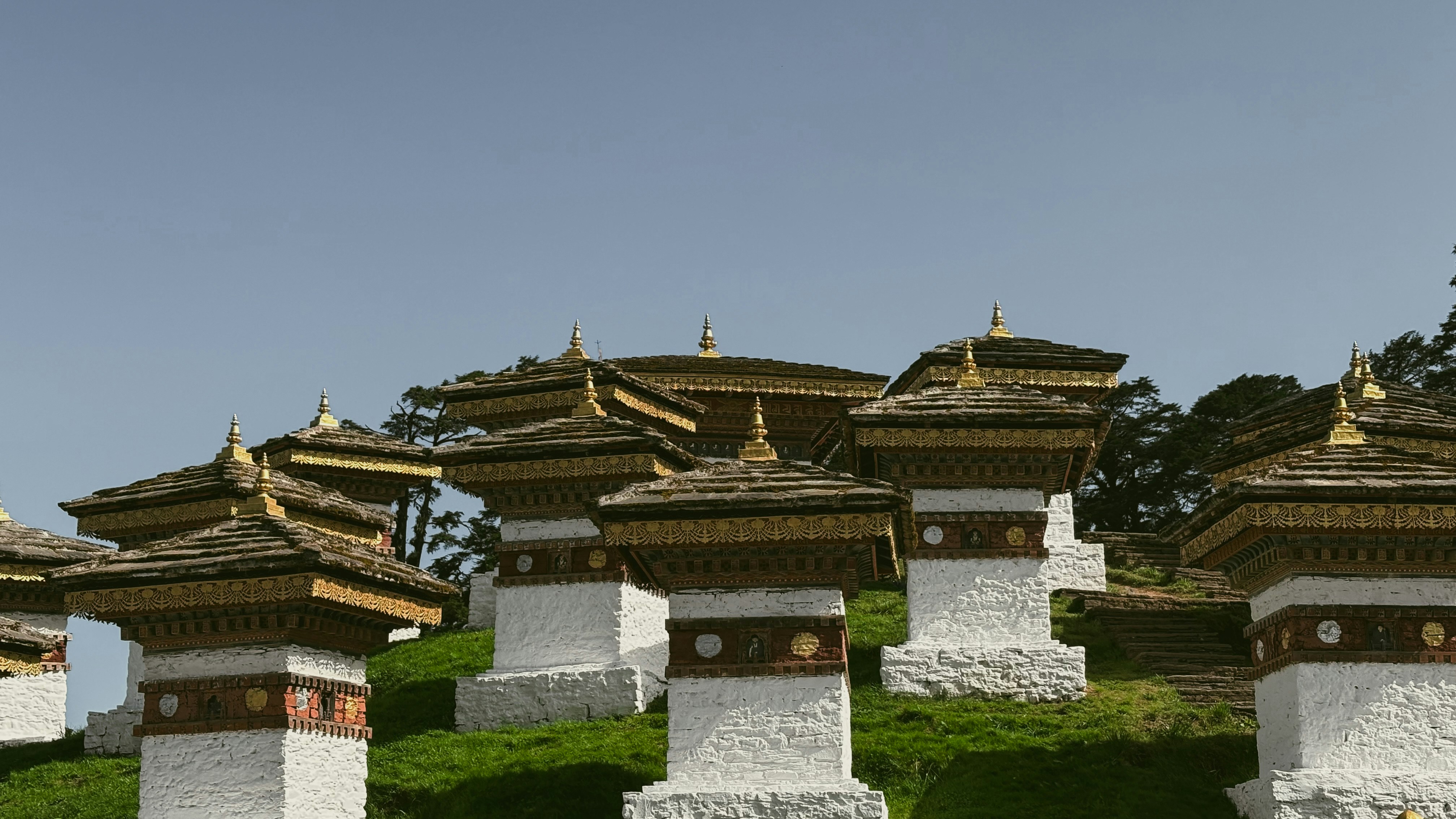 A row of the 108 Druk Wangyal Chortens at Dochula Pass glows under the sunlight, their white walls and golden roofs set against Bhutan’s tranquil mountain backdrop. | White stupas with ornate roofs on a grassy hill.