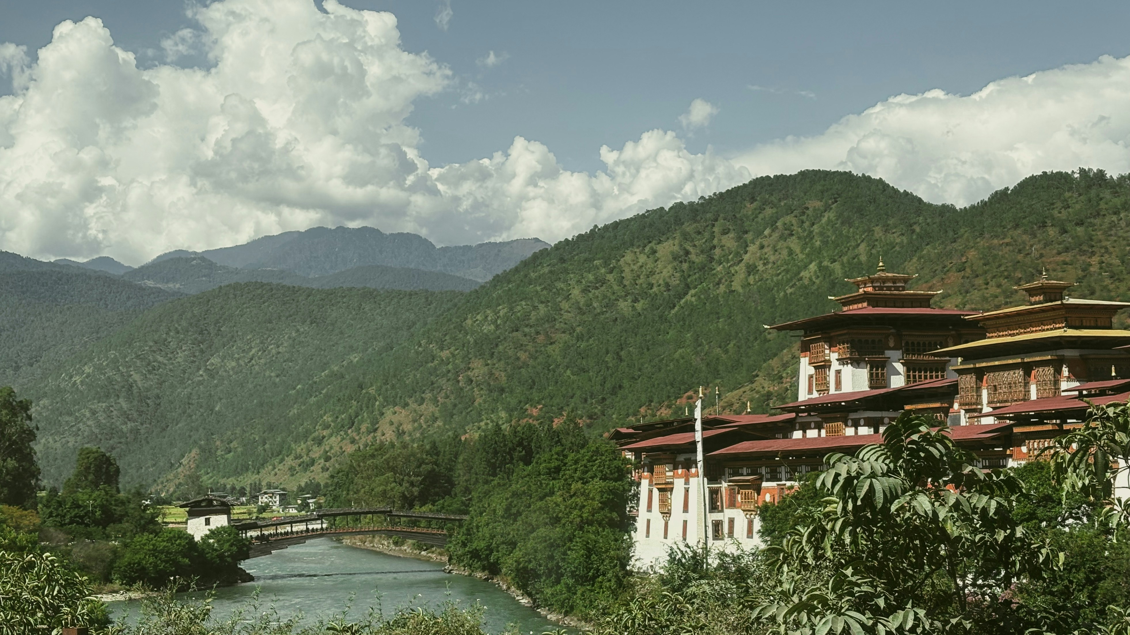 A traditional wooden bridge leads to Paro Dzong, spanning a gentle river beneath the soft glow of dusk, with distant Himalayan peaks completing the tranquil scene.