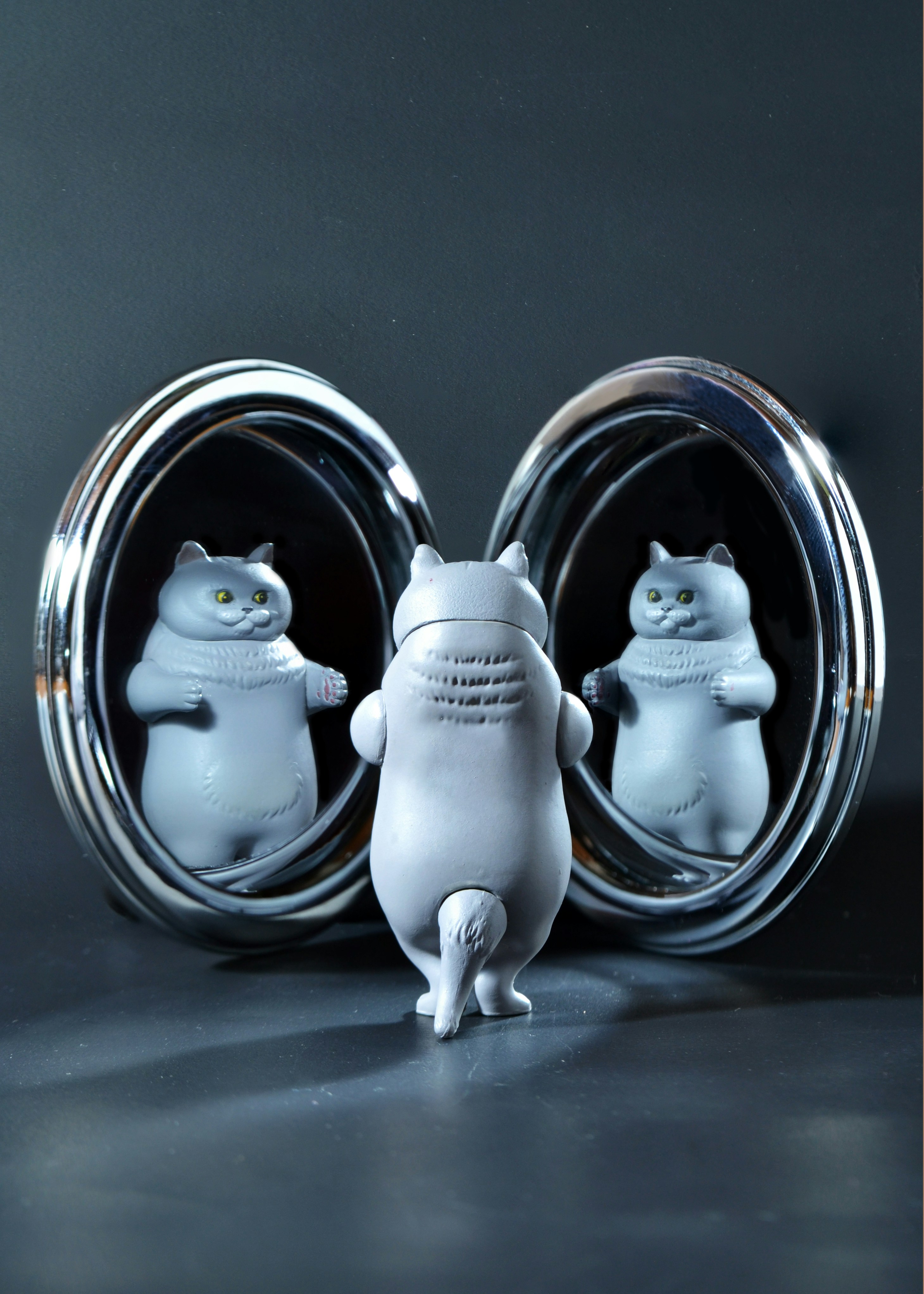 A toy cat and its reflections on the mirror. | Gray cat figurine looking into two mirrors