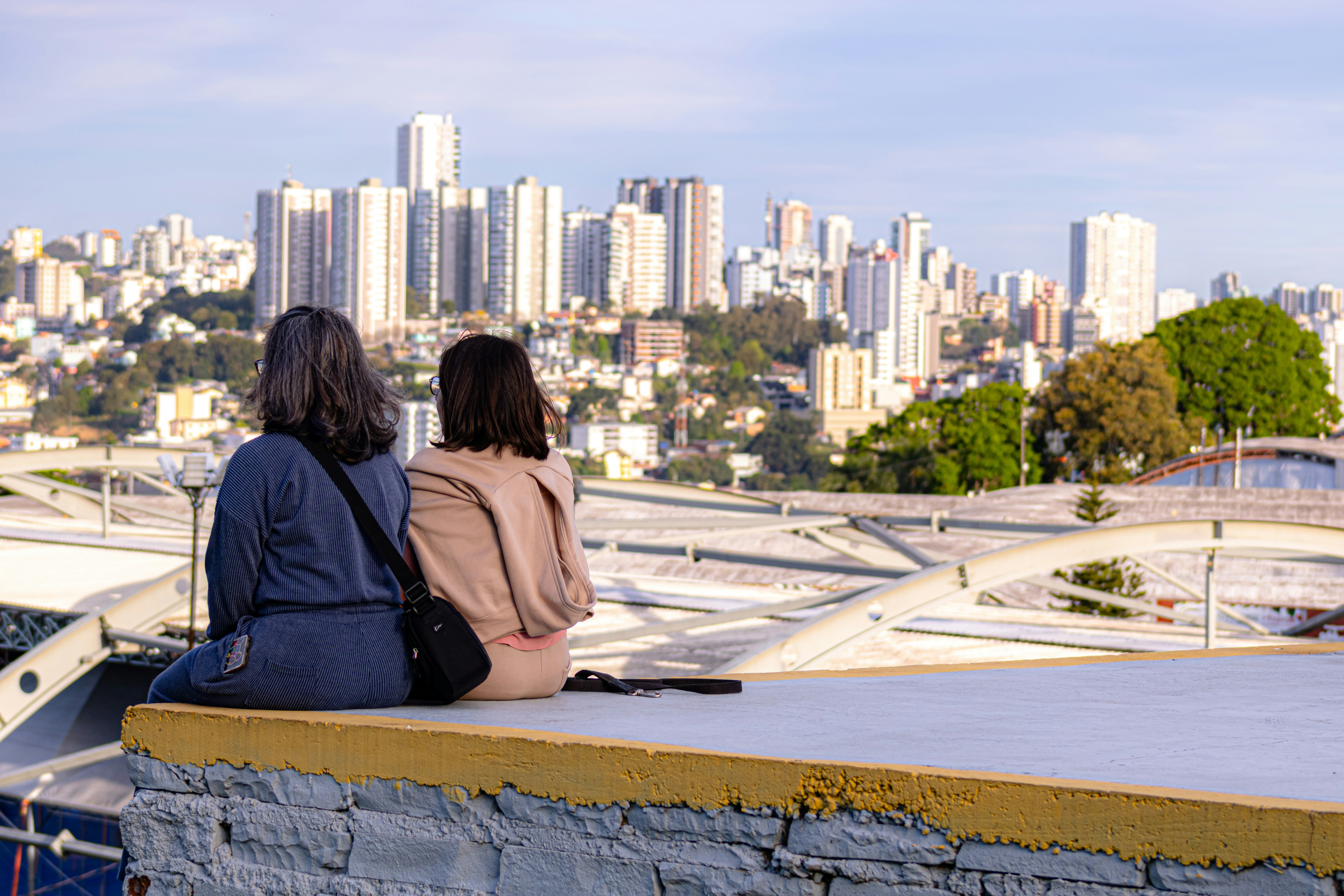 Two women seated on a ledge, gazing over a bustling city skyline filled with modern architecture and greenery.