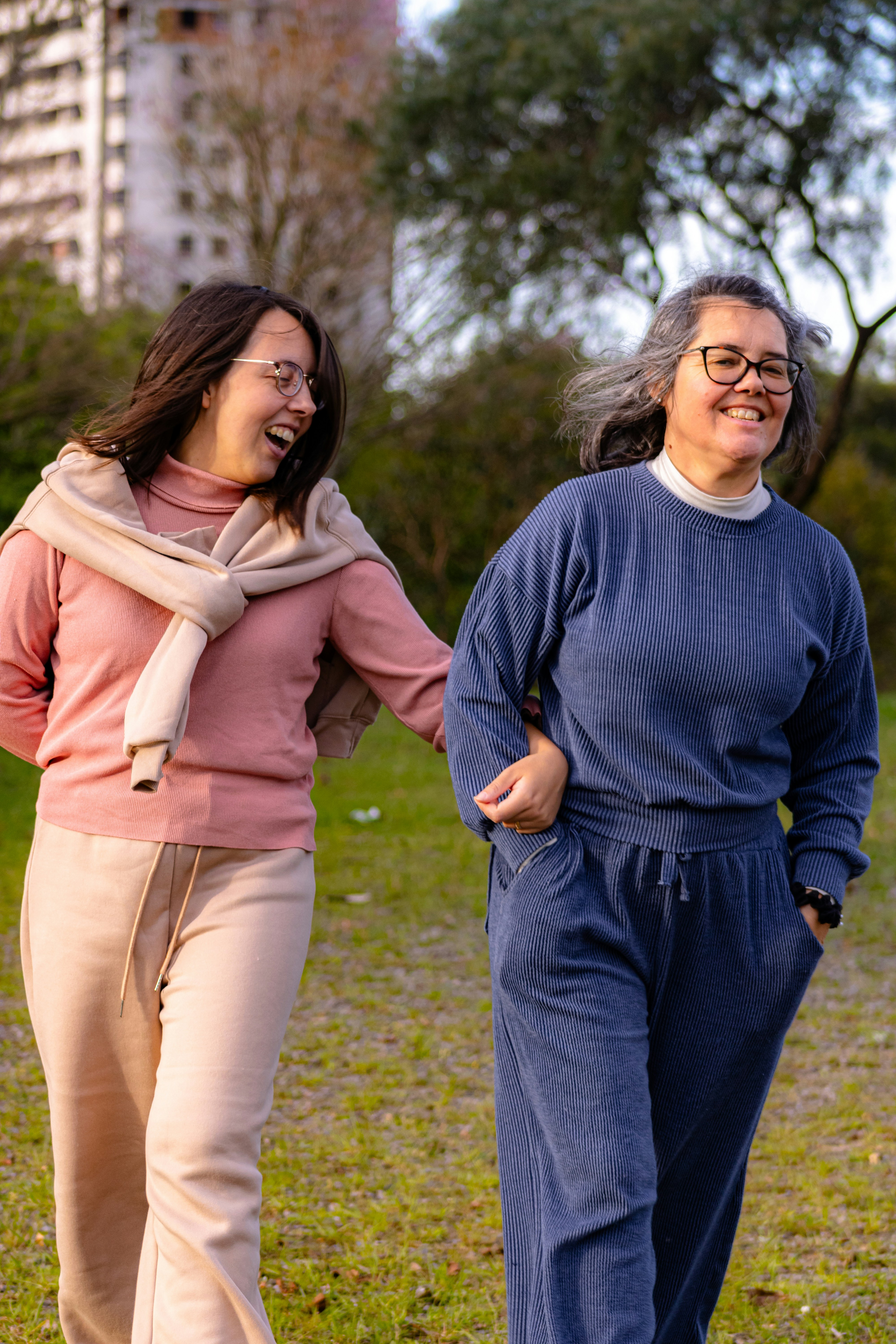 Two friends laughing and enjoying a leisurely walk in a green park with urban buildings in the background.