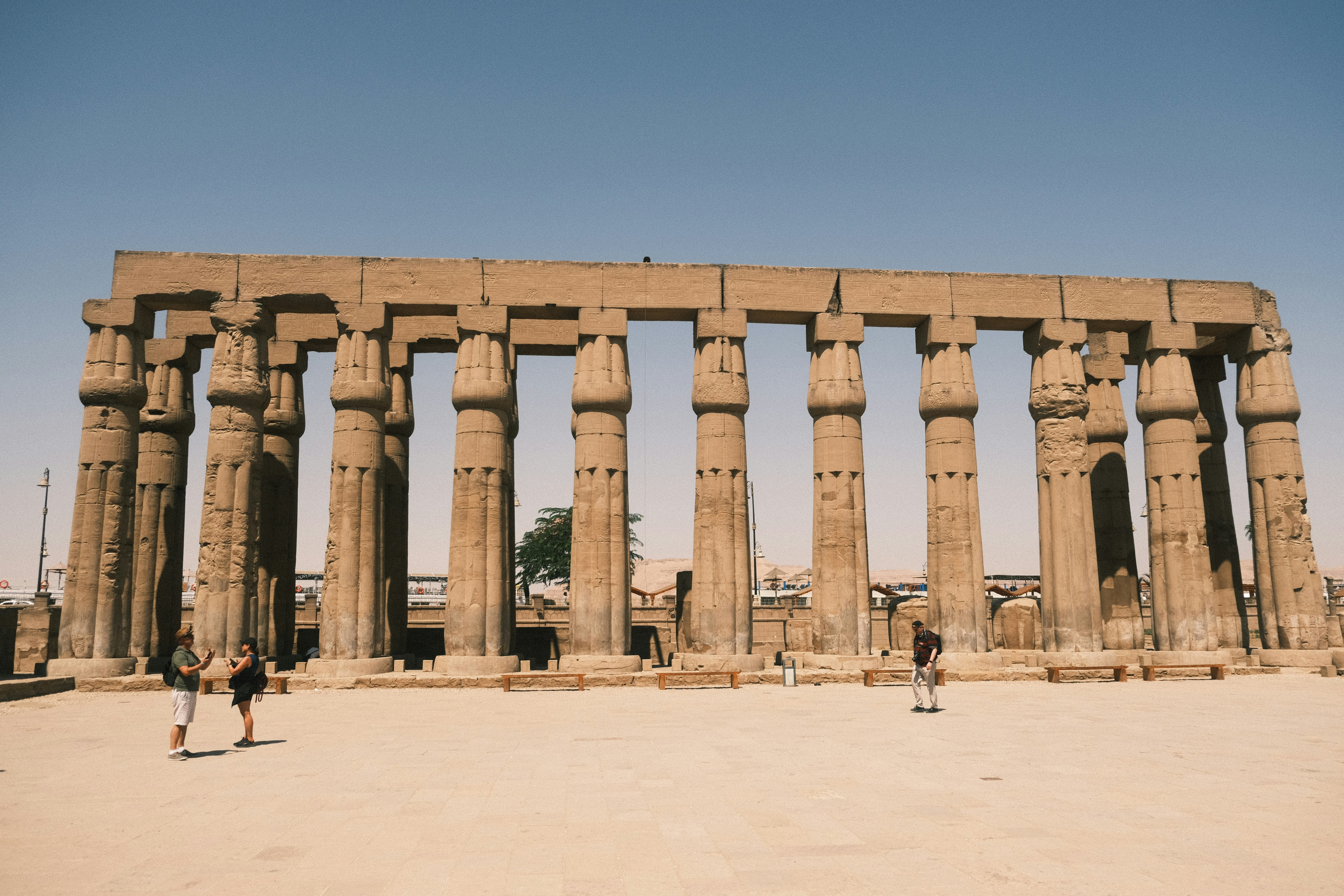 Massive stone pillars of the Luxor Temple rise majestically against a clear blue sky, showcasing intricate carvings. Visitors explore the historic site, immersing themselves in its rich heritage.