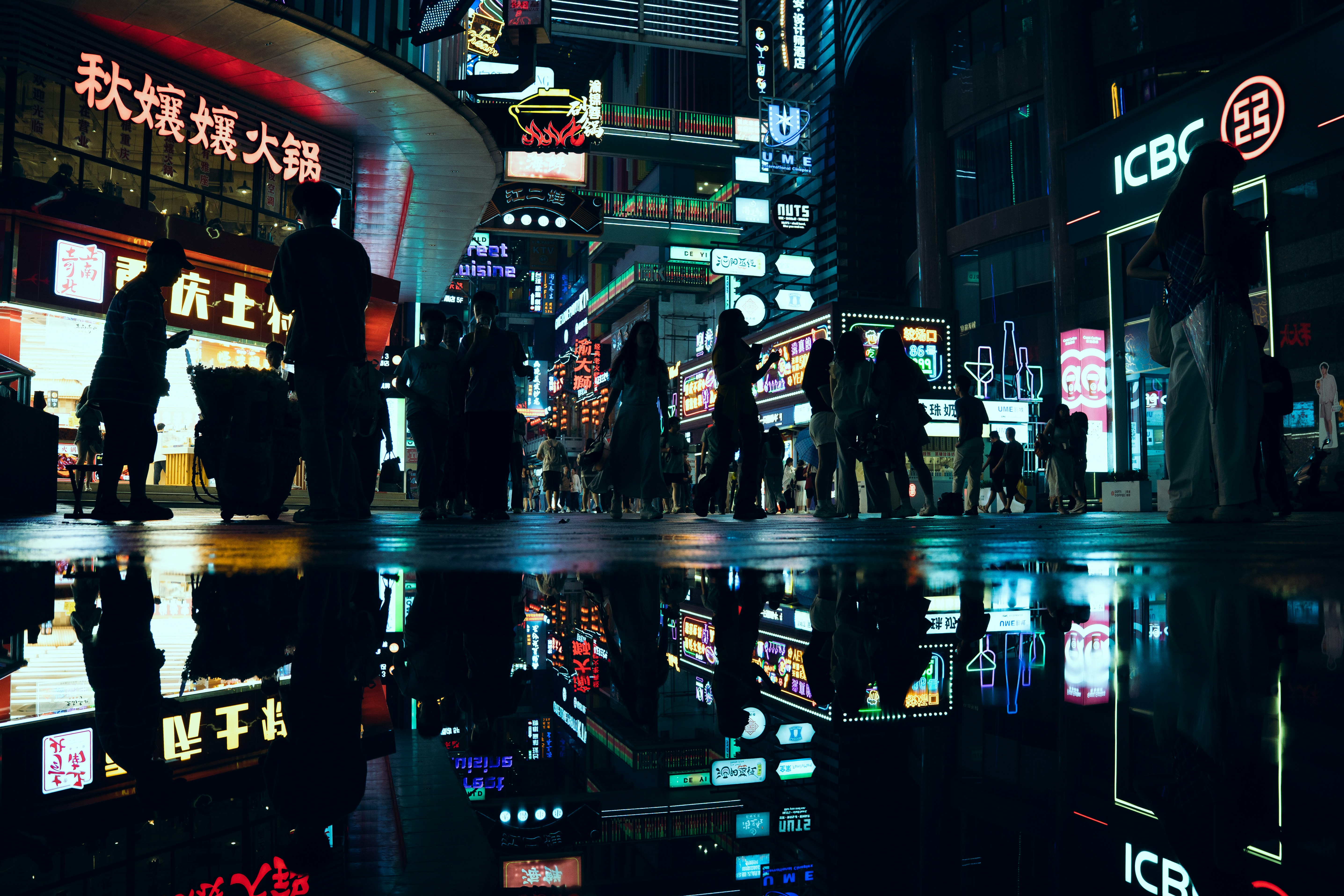 Crowd of silhouettes walking through a vibrant city street, with neon signs reflecting in a puddle on the ground.
