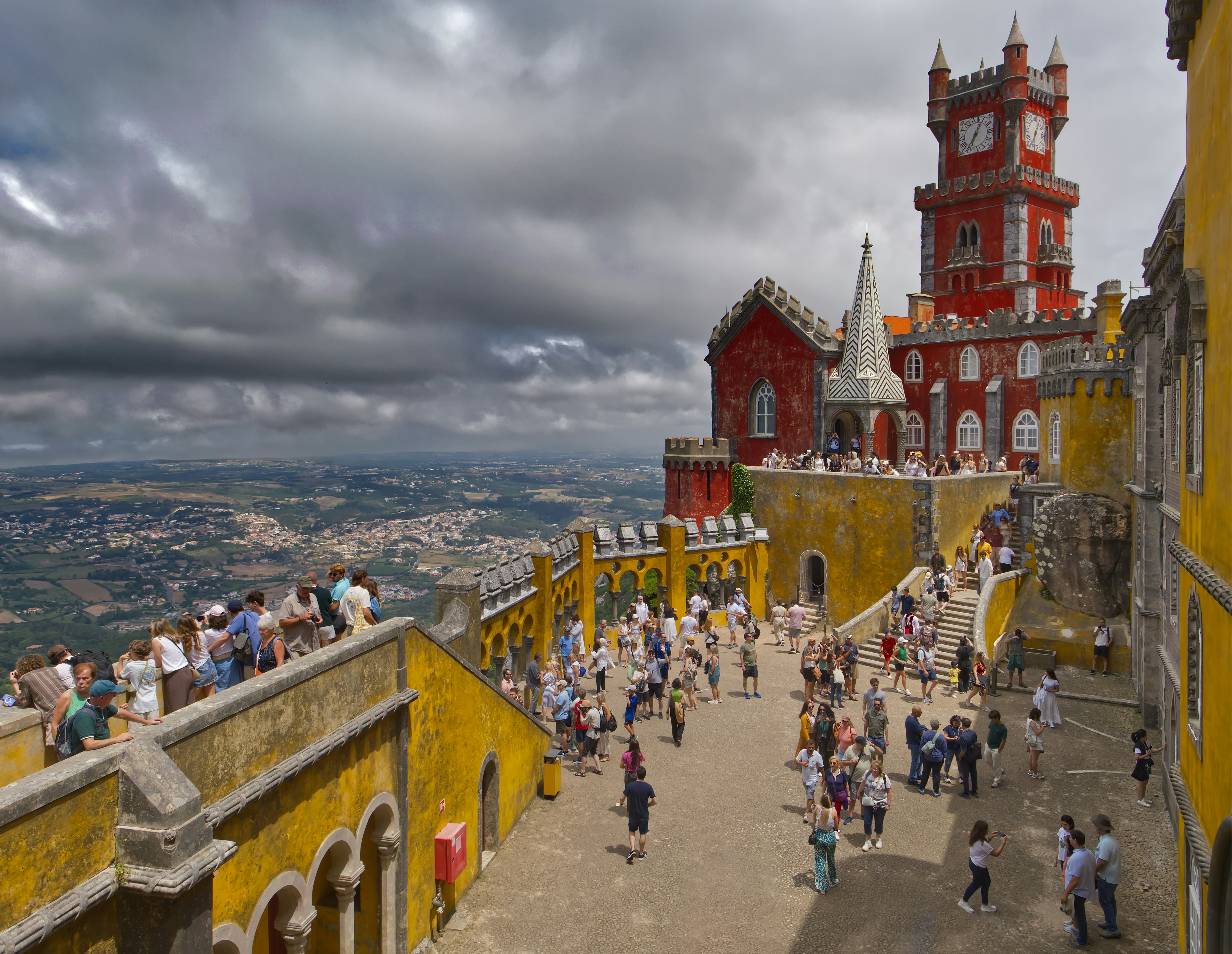 Nous atteignons le point culminant du Palais de Pena. D’ici, les chemins de ronde dévoilent un panorama à couper le souffle sur la campagne de Sintra, qui s’étend à perte de vue sous un ciel changeant. En contrebas, la foule s’anime — un véritable ballet de visiteurs venus des quatre coins du monde, tous captivés par cette œuvre d’architecture fantasque, où s’entremêlent les influences mauresques, gothiques et renaissantes. | Colorful palace on a hill with many tourists