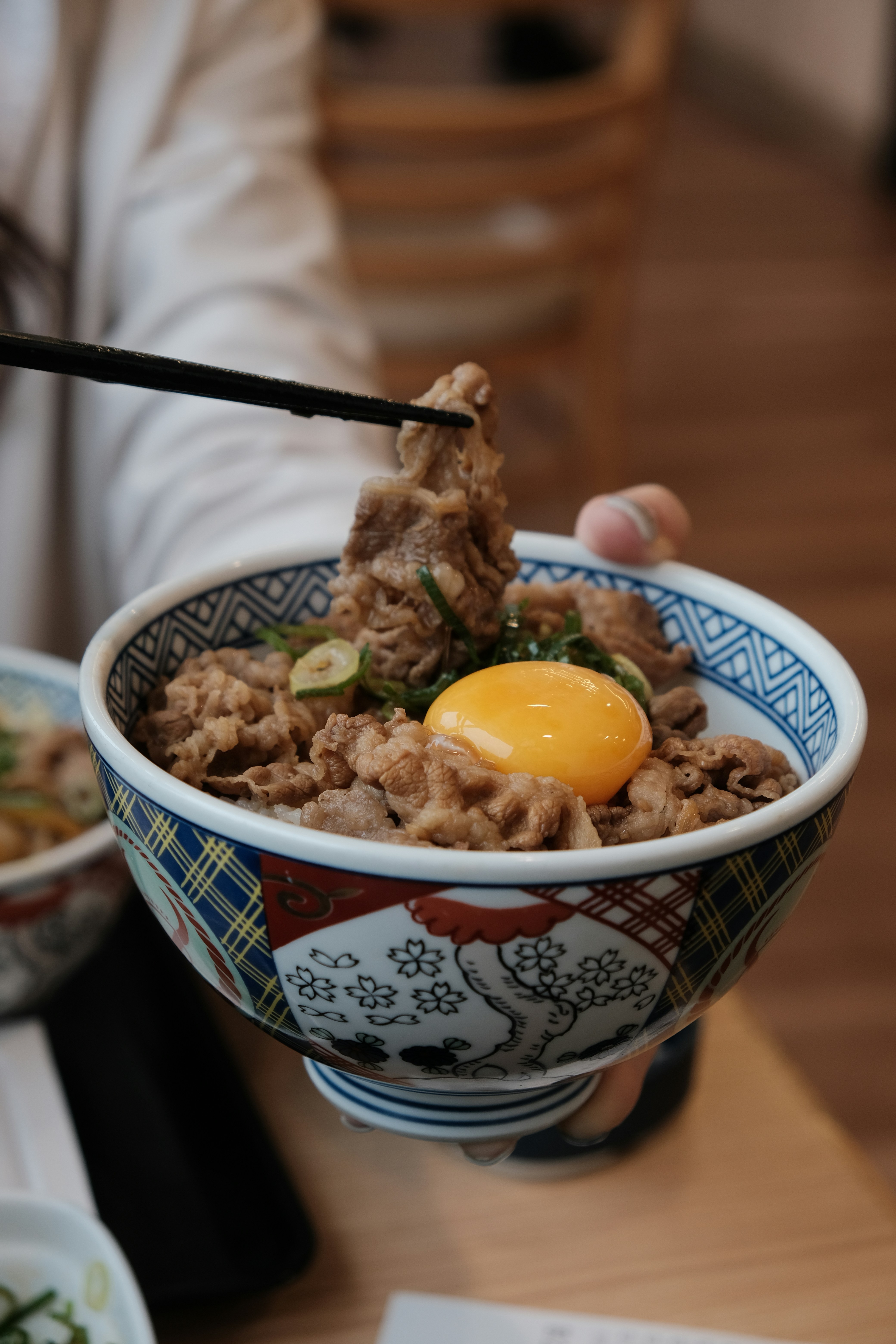 A hand holding a bowl of gyudon topped with a raw egg, showcasing tender beef and green onions. The intricate bowl design adds cultural flair.
