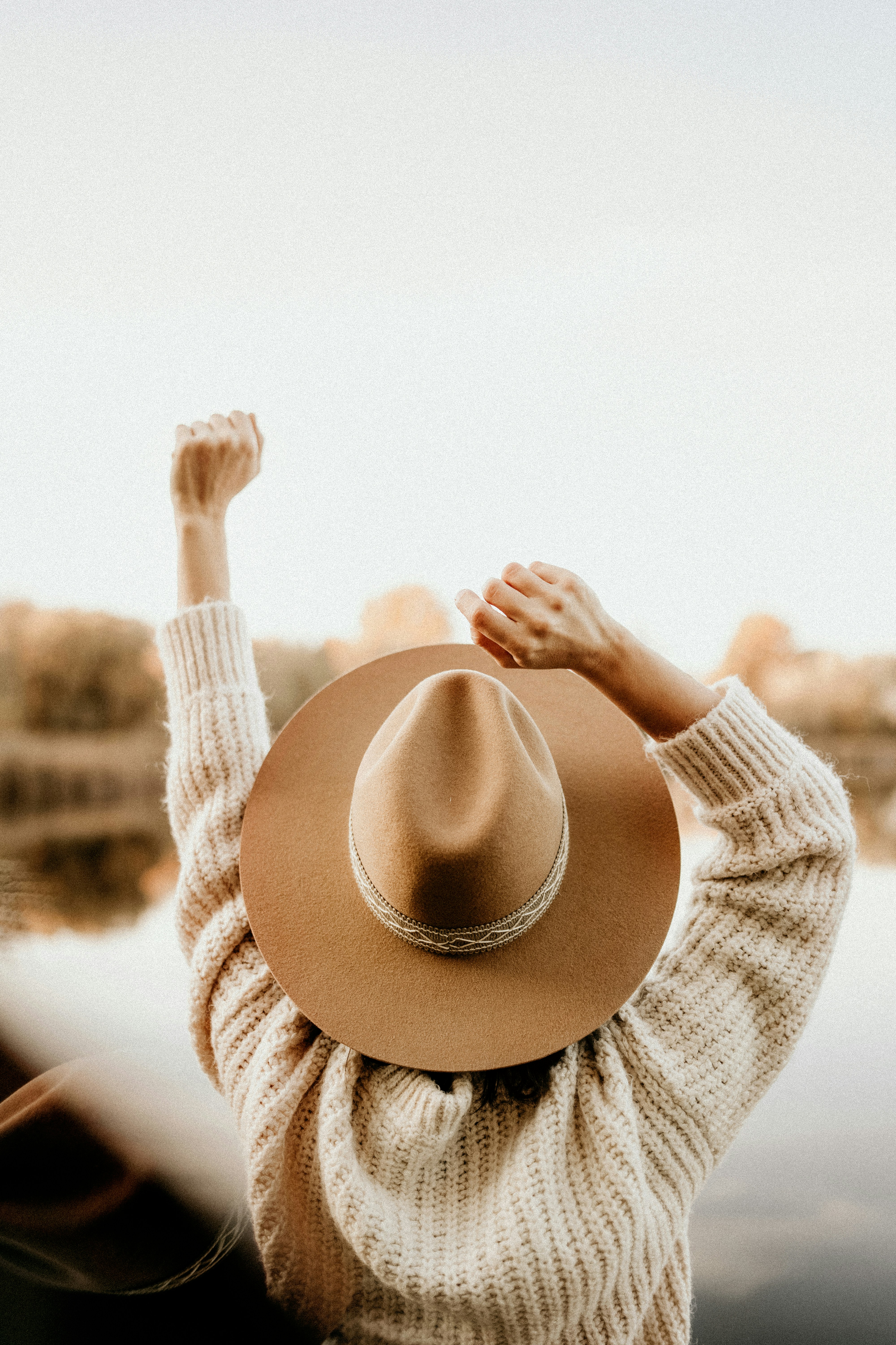 Woman in hat raises arms in celebration by water