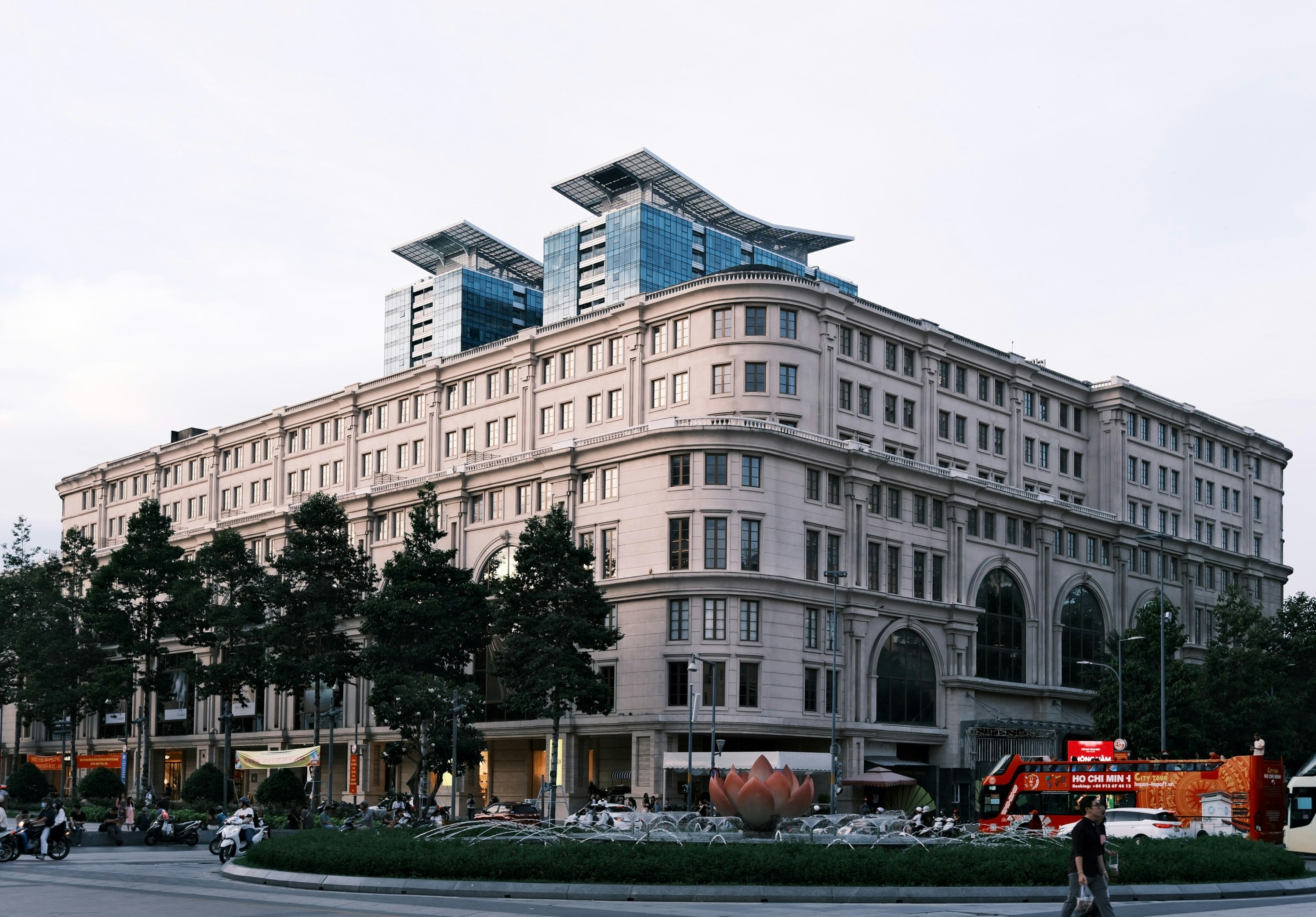 Historic building with modern glass structures atop, surrounded by lush greenery and urban life.