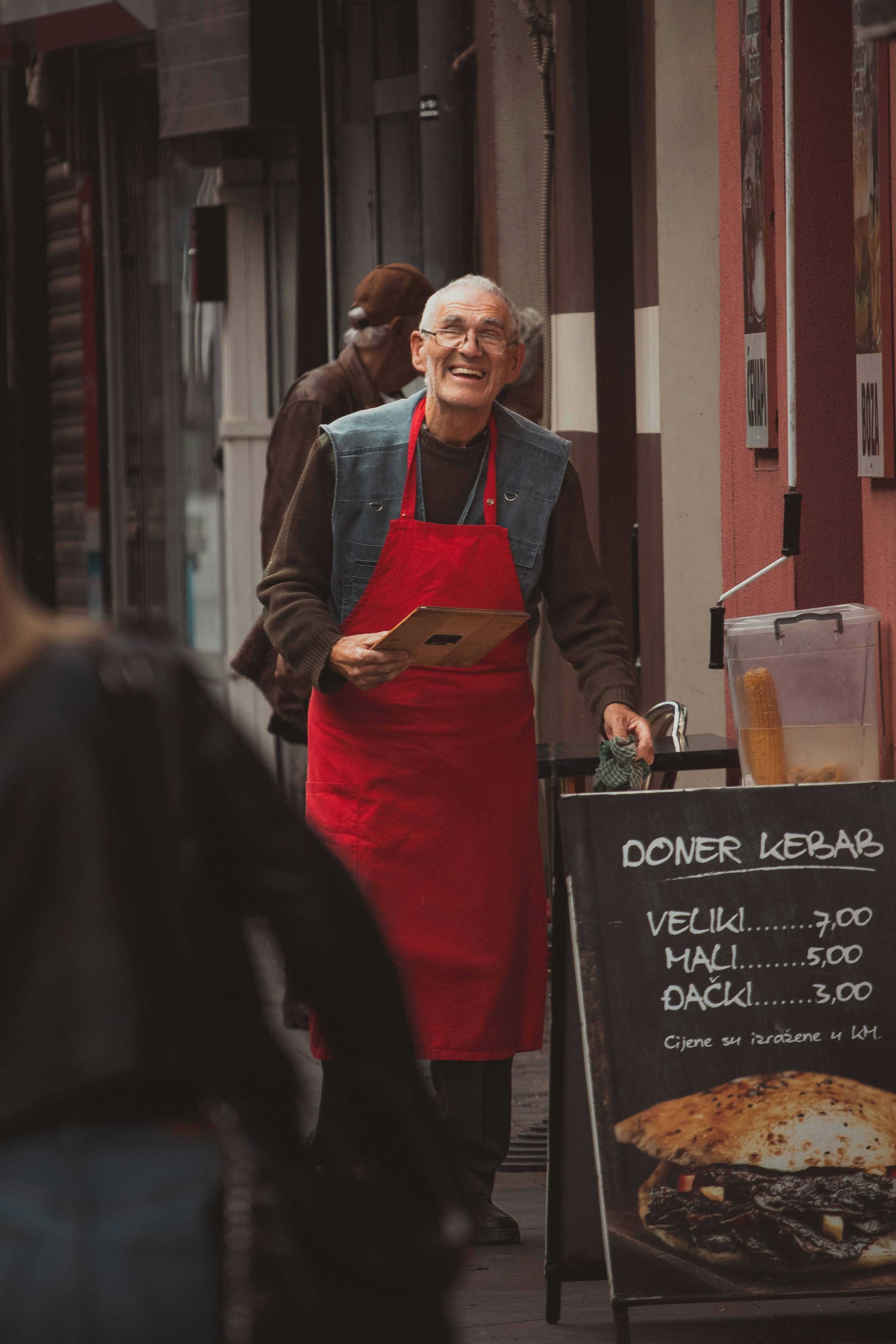A cheerful vendor in a red apron stands by his food cart, offering delicious döner kebabs to passersby. The vibrant street scene captures the essence of local culinary culture.