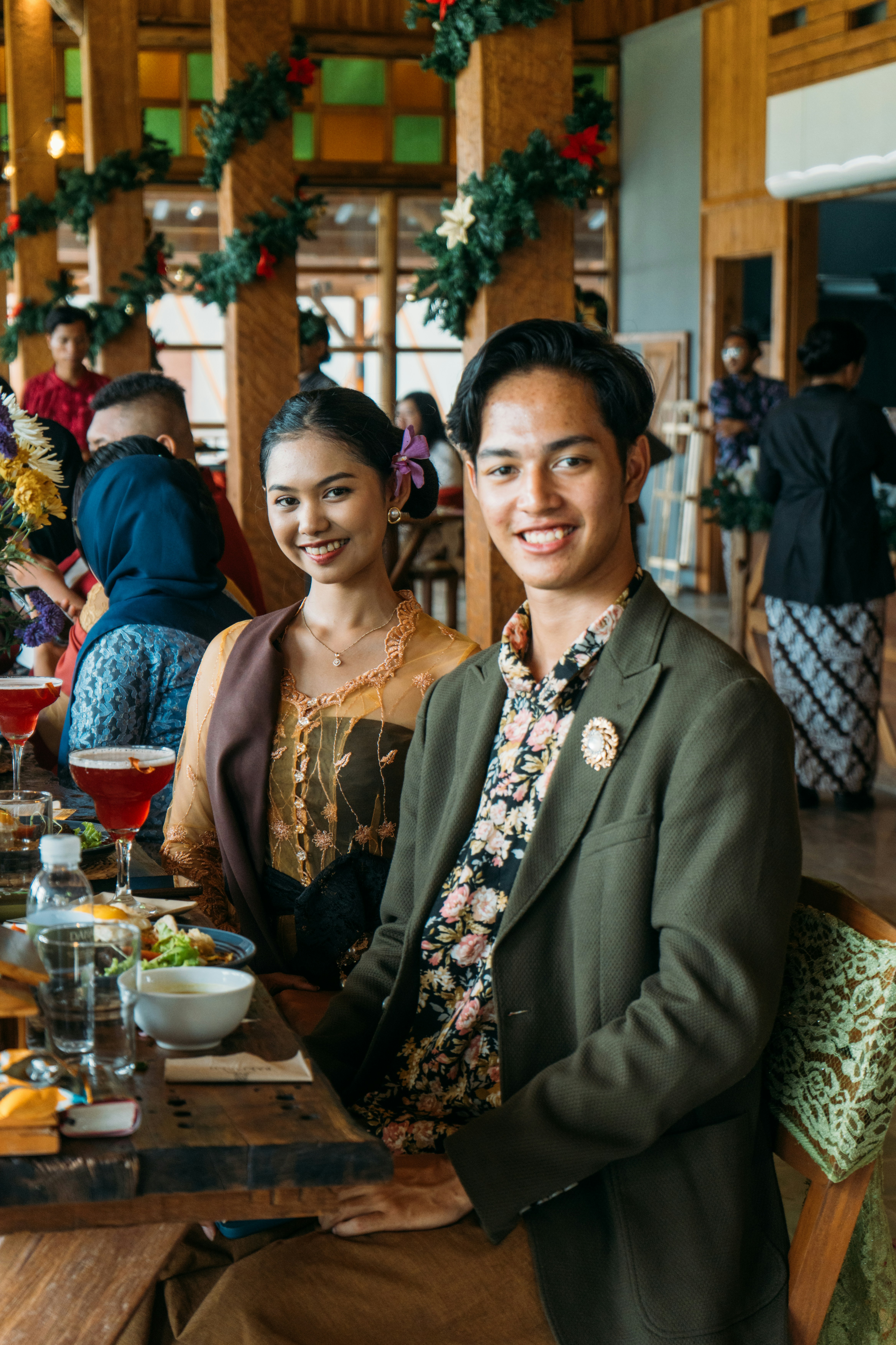 A young couple dressed in traditional attire smiles at the camera during a festive gathering, surrounded by elegantly arranged food and drinks.