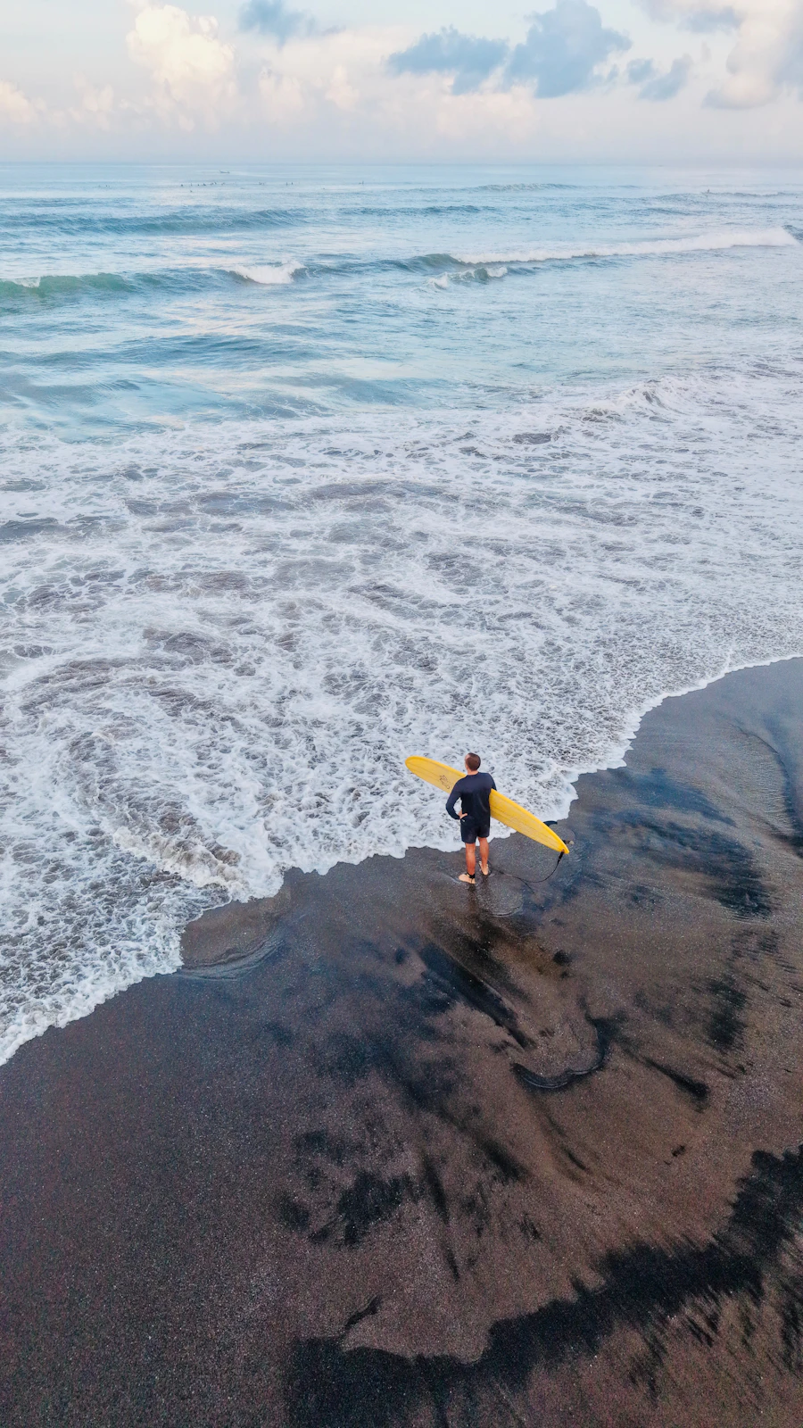Surfer standing on the shoreline with a yellow surfboard, gazing at the ocean waves. The contrast of the dark sand and foamy water creates a dynamic scene.