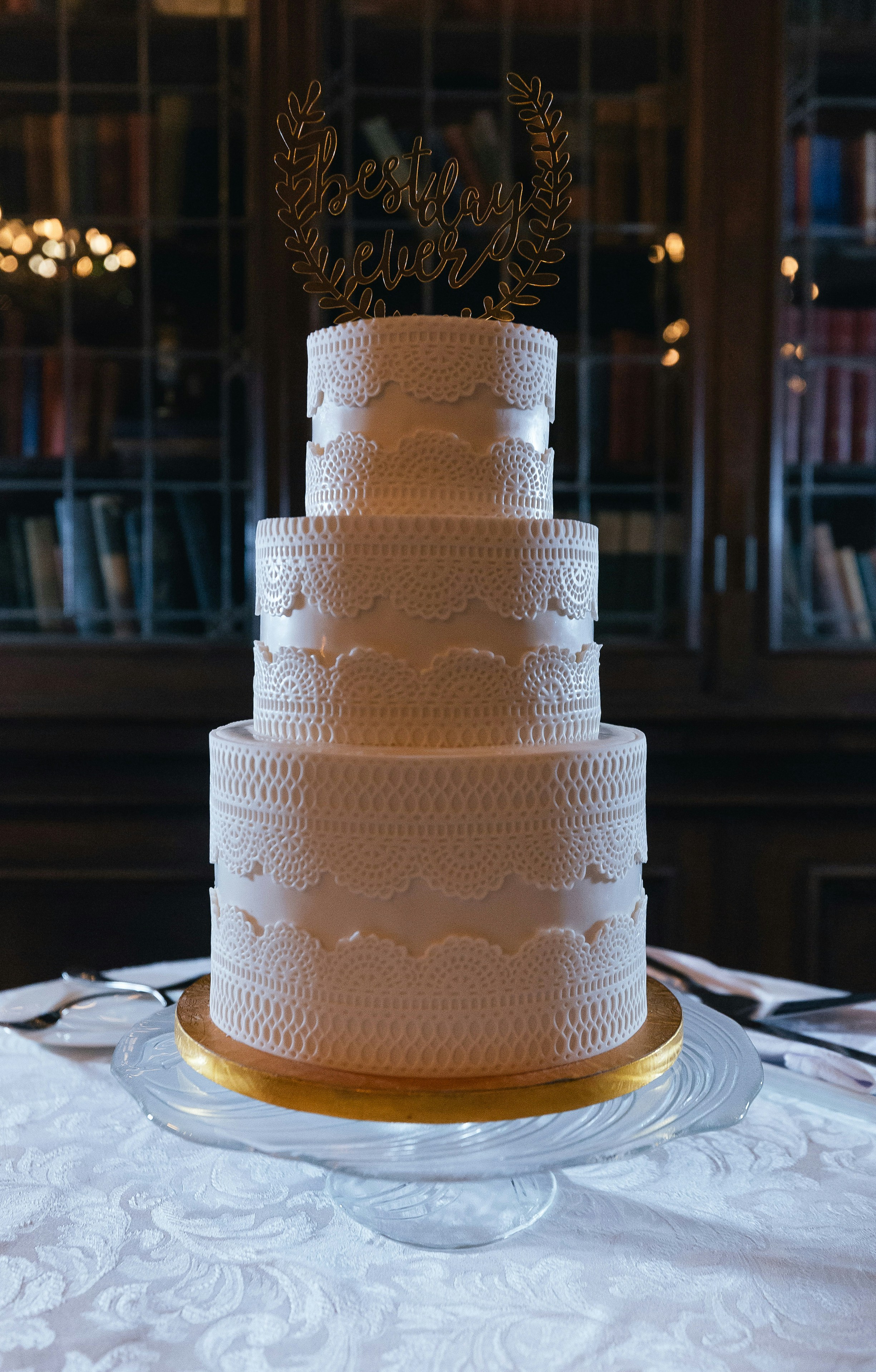 Three-tiered wedding cake adorned with intricate lace patterns and a golden topper proclaiming 'best day ever.'