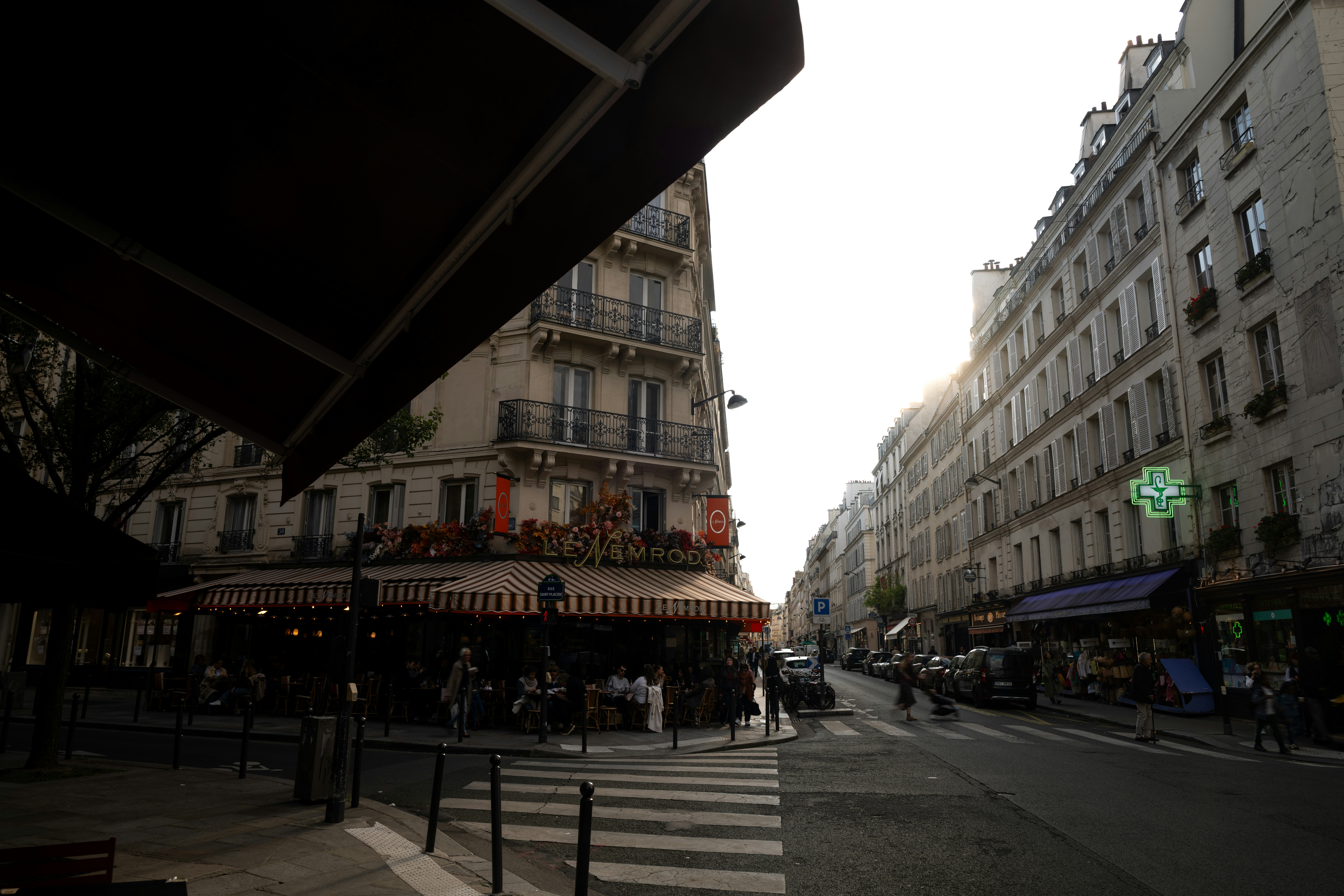 Bustling Parisian café at the corner of a busy street, with patrons enjoying their time under the striped awning. The warm glow of the sun adds a nostalgic touch.