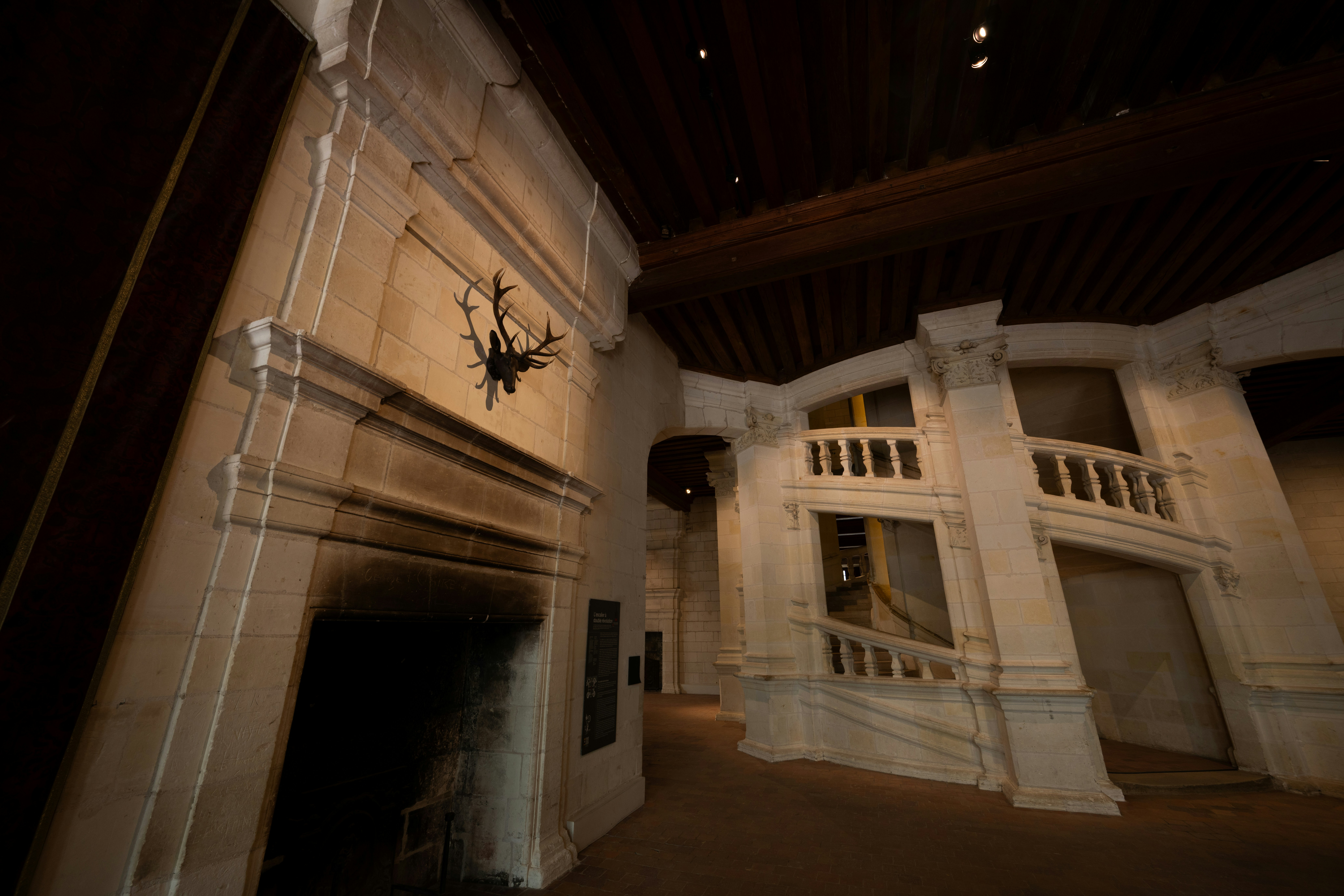 Intricate stone fireplace adorned with a decorative antler mount, leading to a grand staircase in a historic interior space.