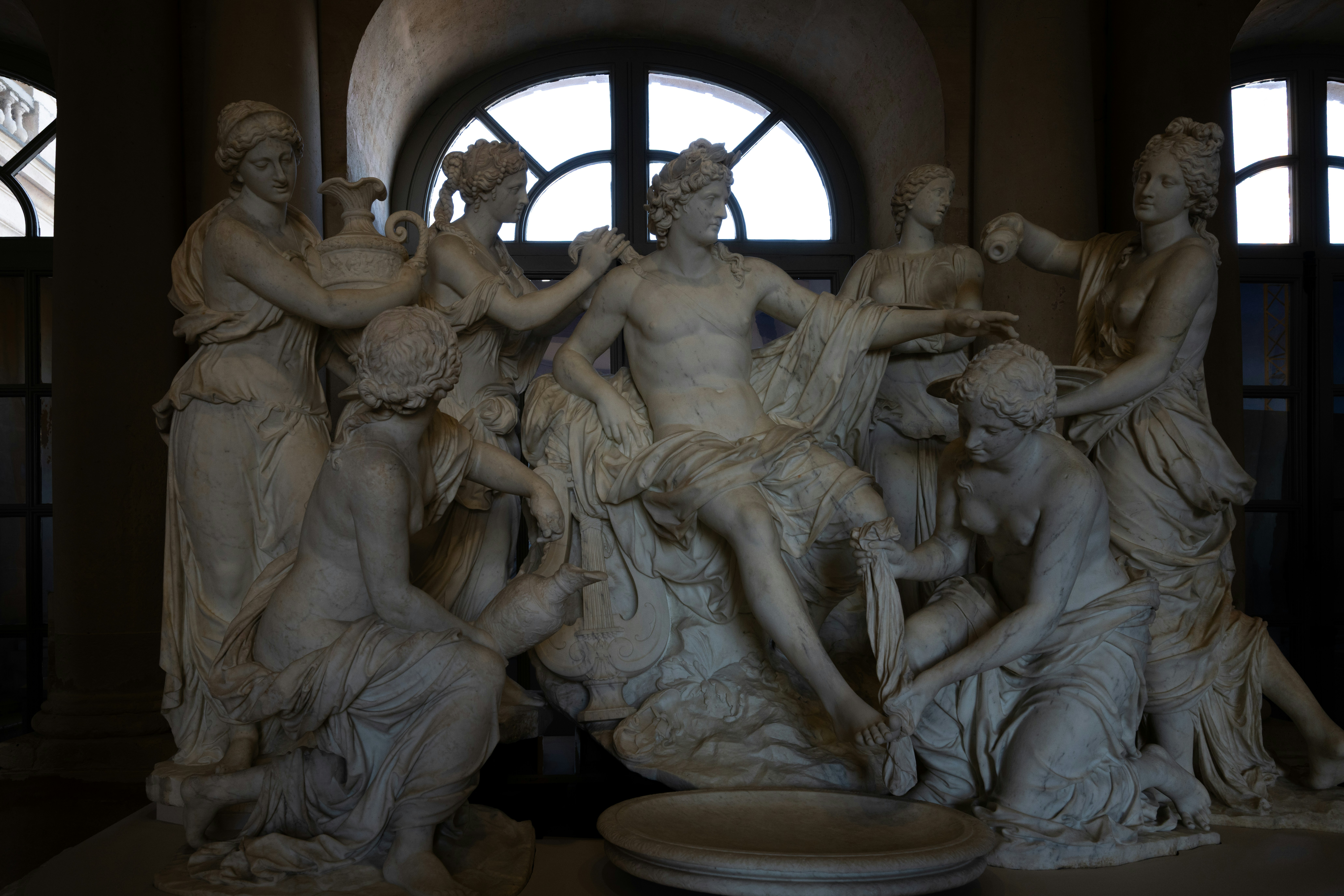 A classical marble sculpture depicting a scene of muses attending to a central figure, showcasing intricate details and expressions. The artwork captures a moment of artistic inspiration.