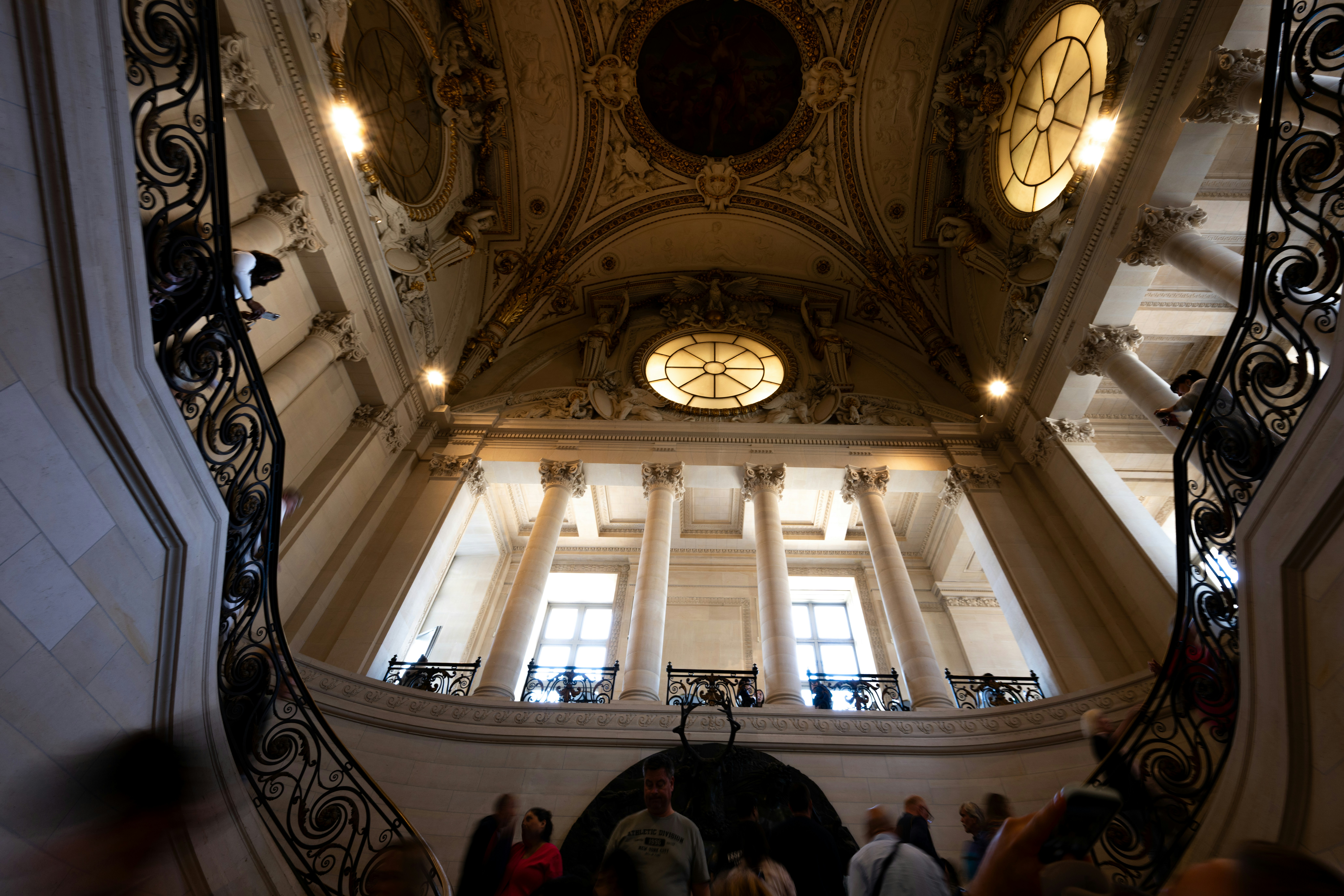 Intricate staircase with ornate railings leading to grand windows, showcasing the architectural beauty of the interior space. People move through the scene, adding a sense of life and dynamism.