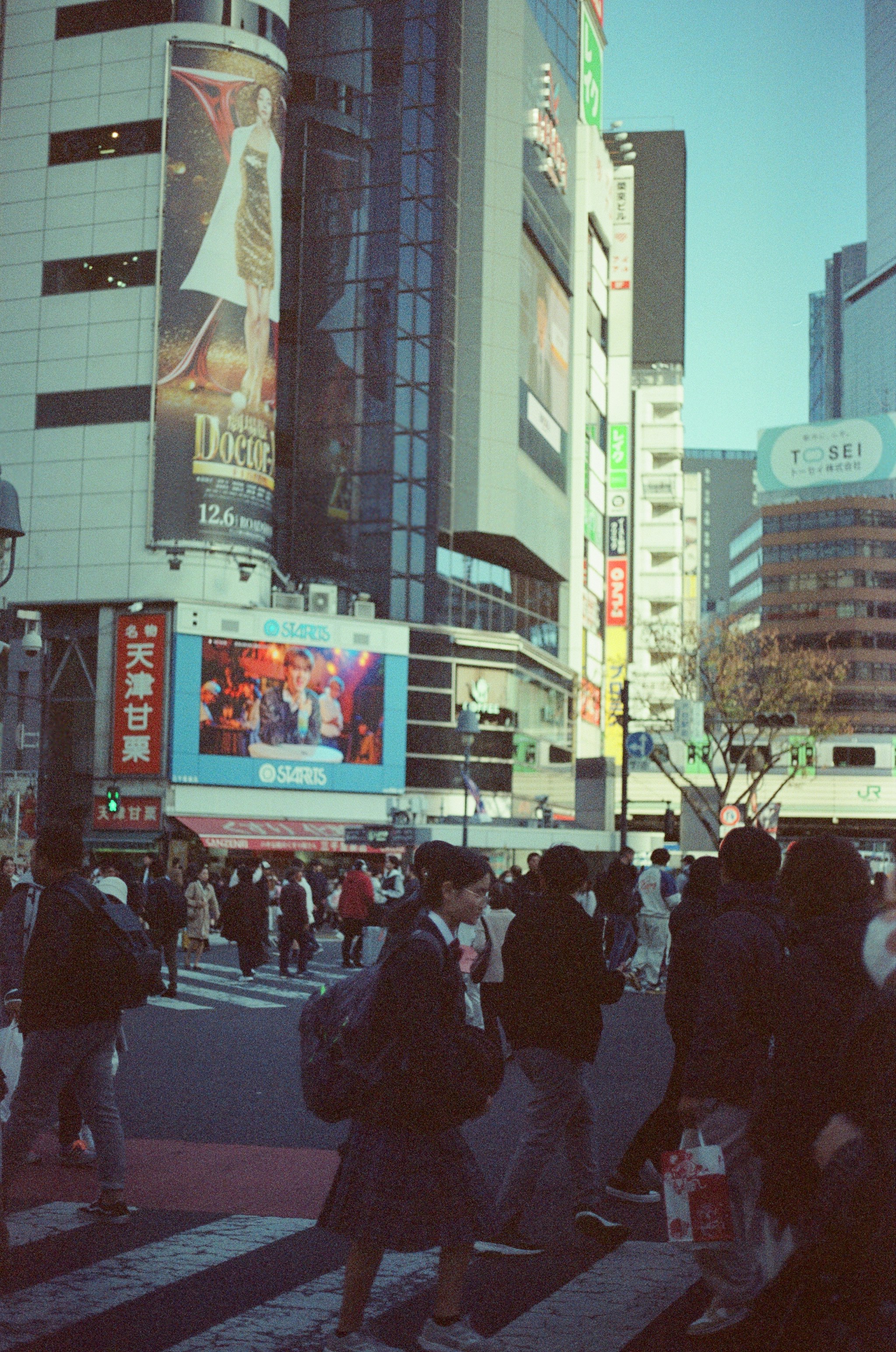 A bustling urban crosswalk filled with pedestrians navigating the vibrant cityscape, featuring large advertisements on surrounding buildings.