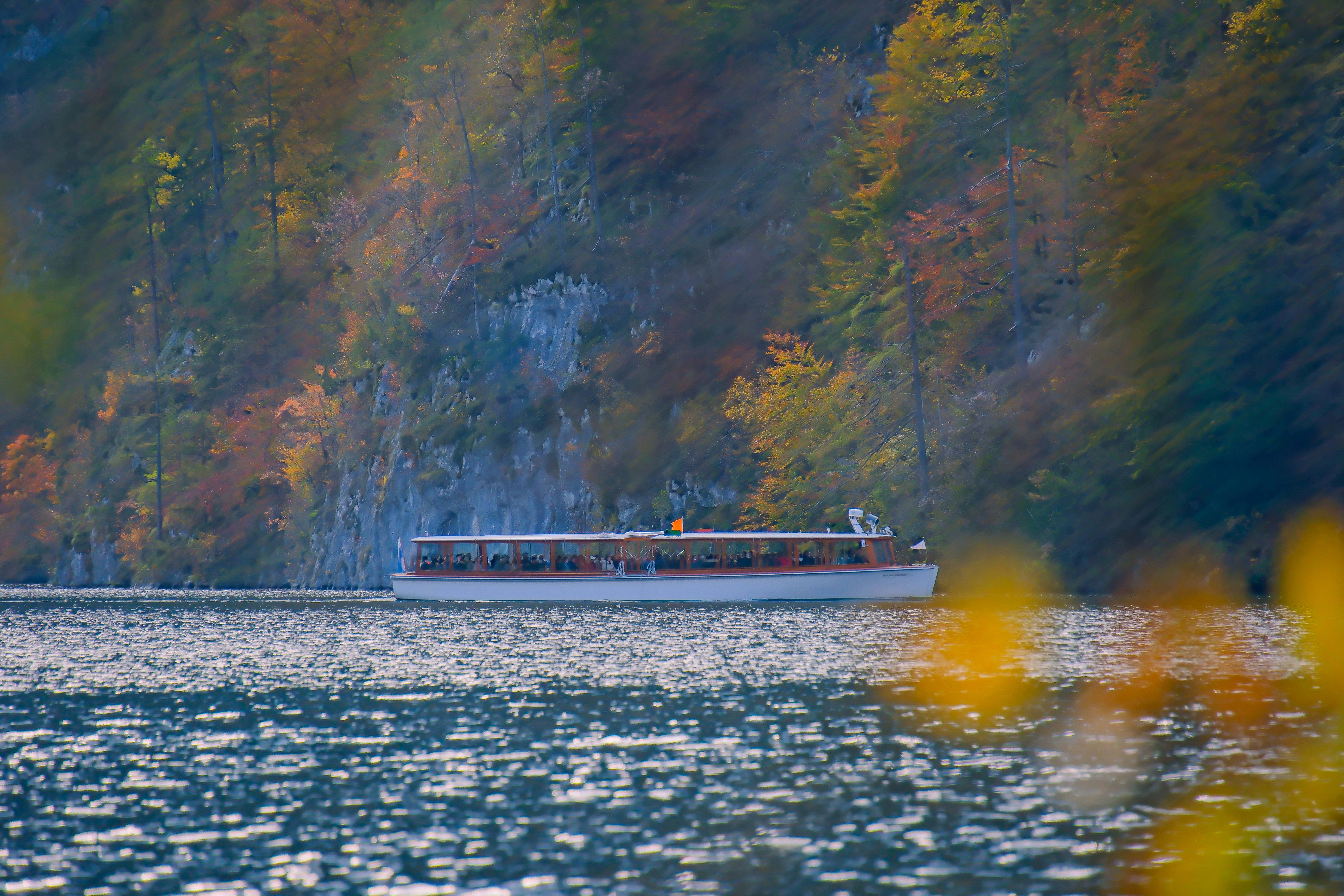 A serene autumn scene at Lake Königssee, with a passenger boat gliding across shimmering water. | Tour boat on a lake with autumn foliage