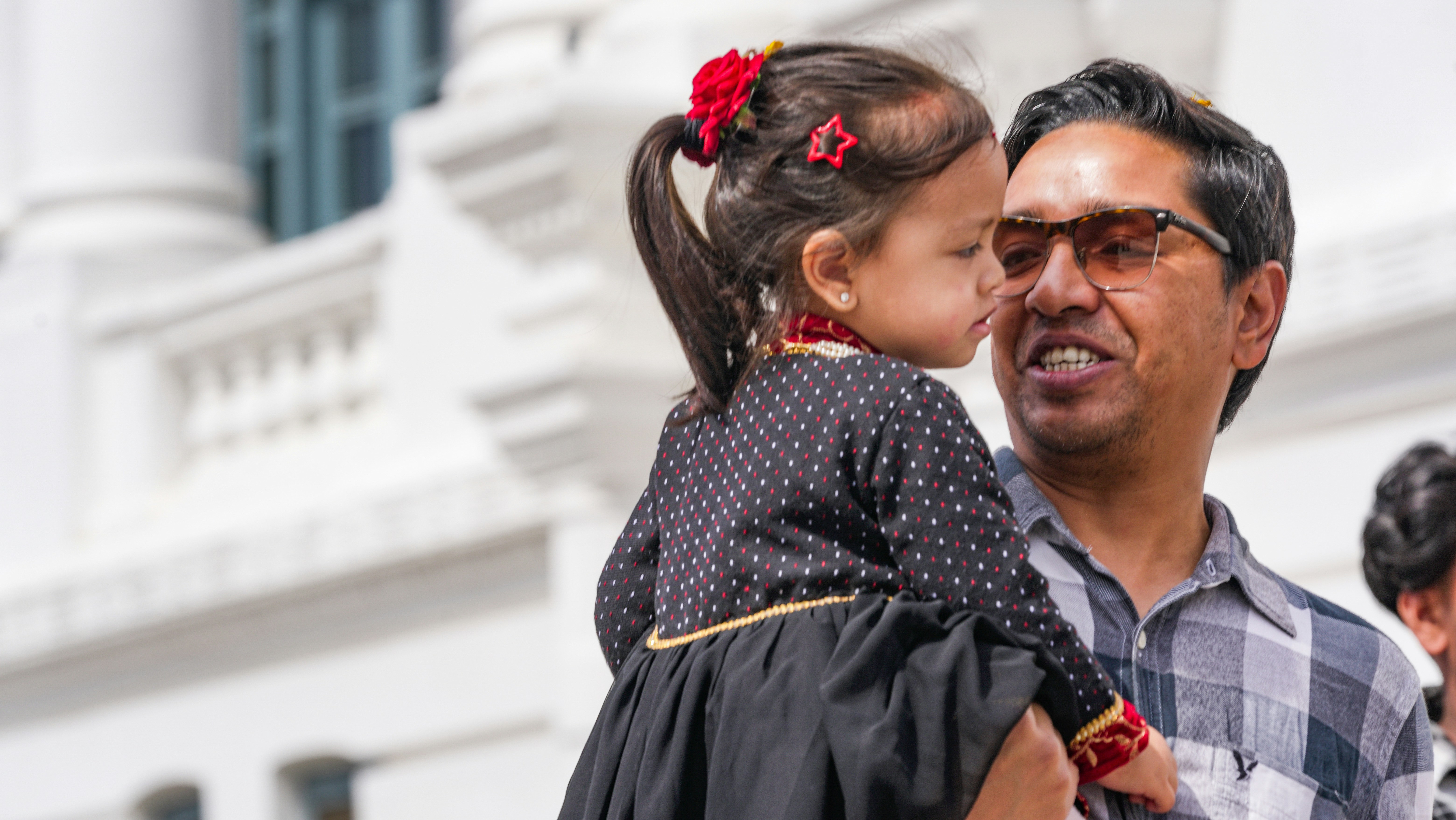 Father holding his daughter, both dressed in traditional attire, sharing a joyful moment outdoors. The background features architectural elements that enhance the cultural setting.