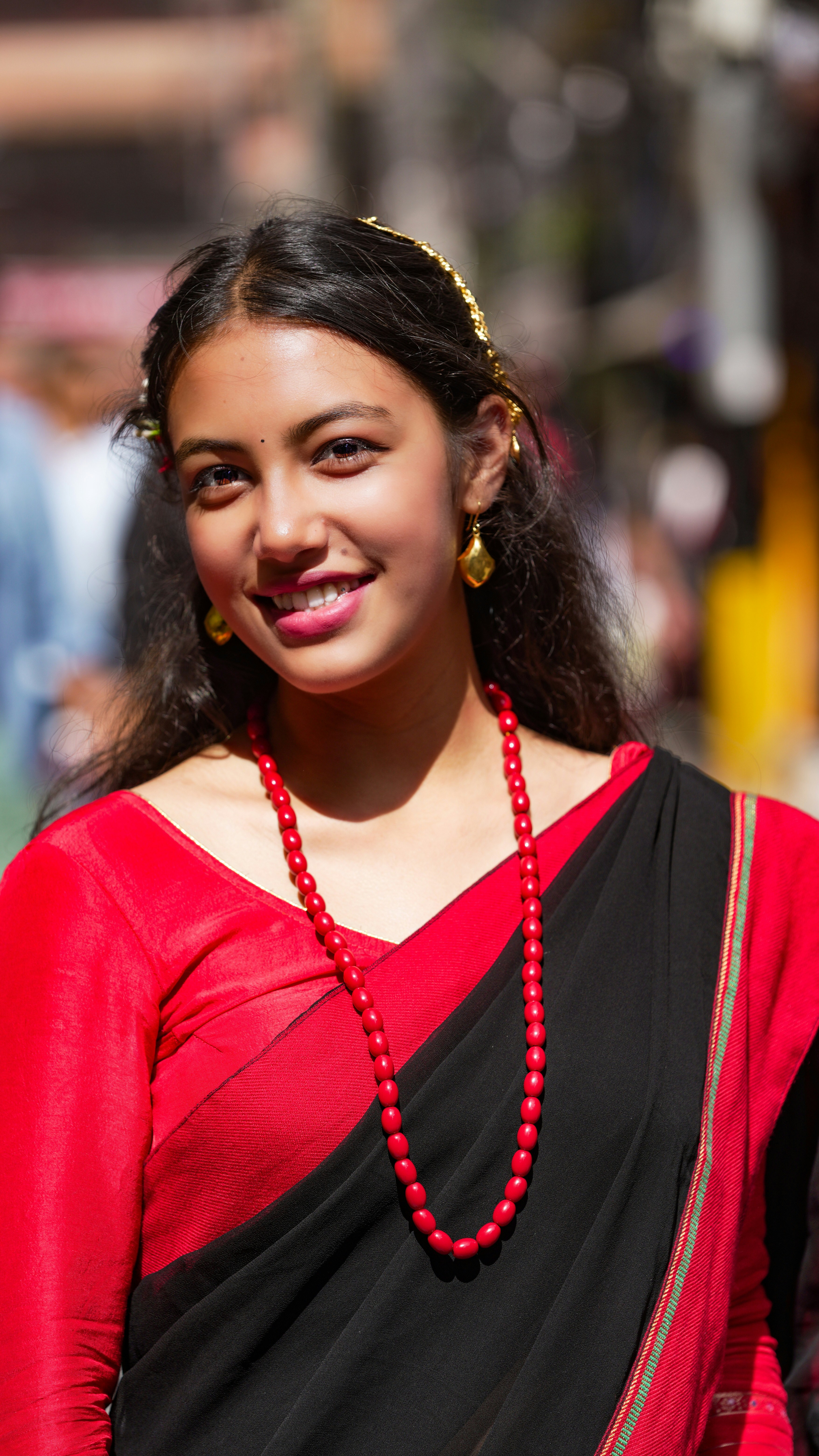 Young woman in a traditional black and red saree adorned with red beads, smiling warmly against a lively street backdrop.