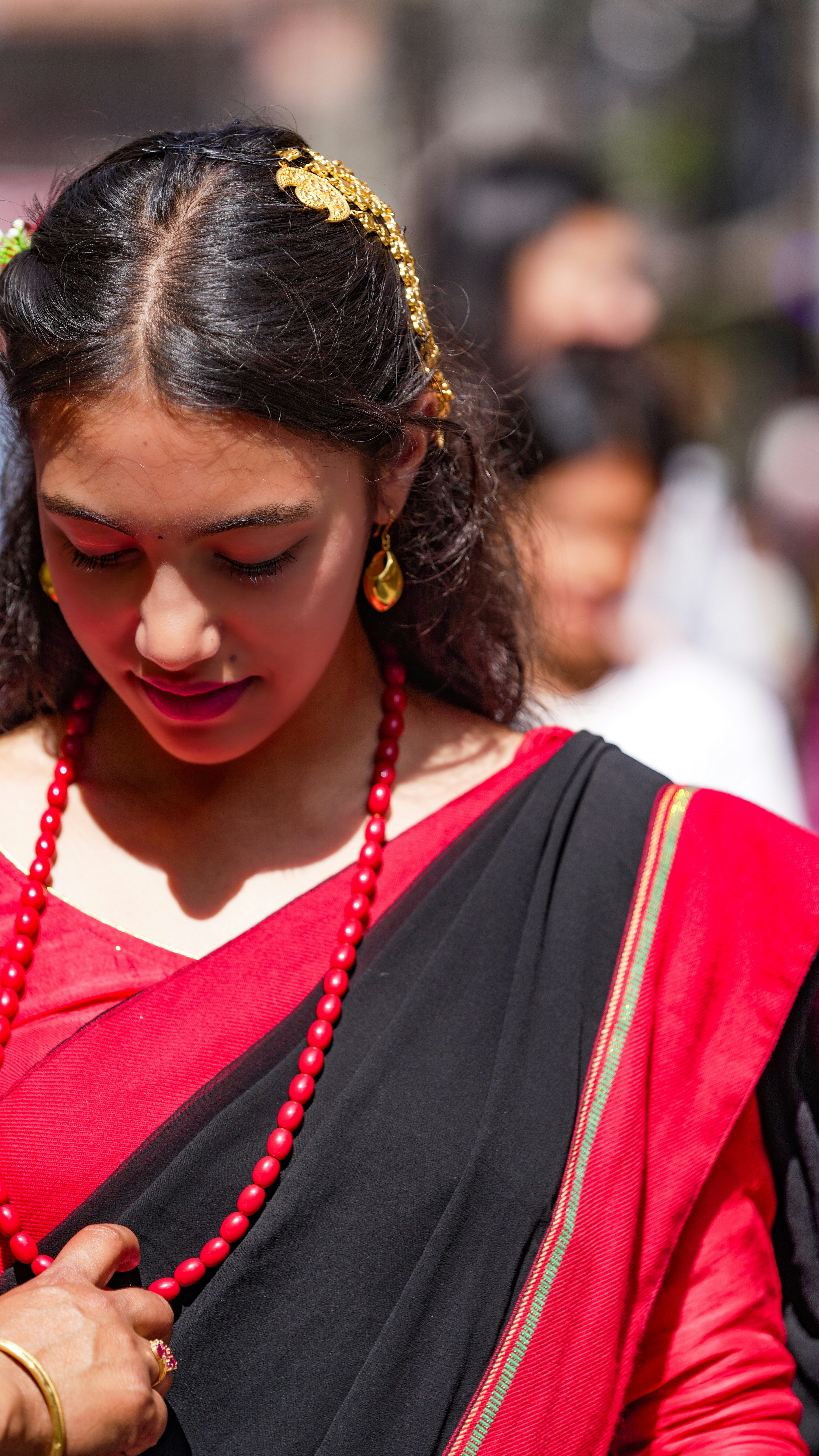 A young woman in a vibrant red and black saree, adorned with jewelry, gazes down thoughtfully as she adjusts her necklace amidst a bustling crowd.