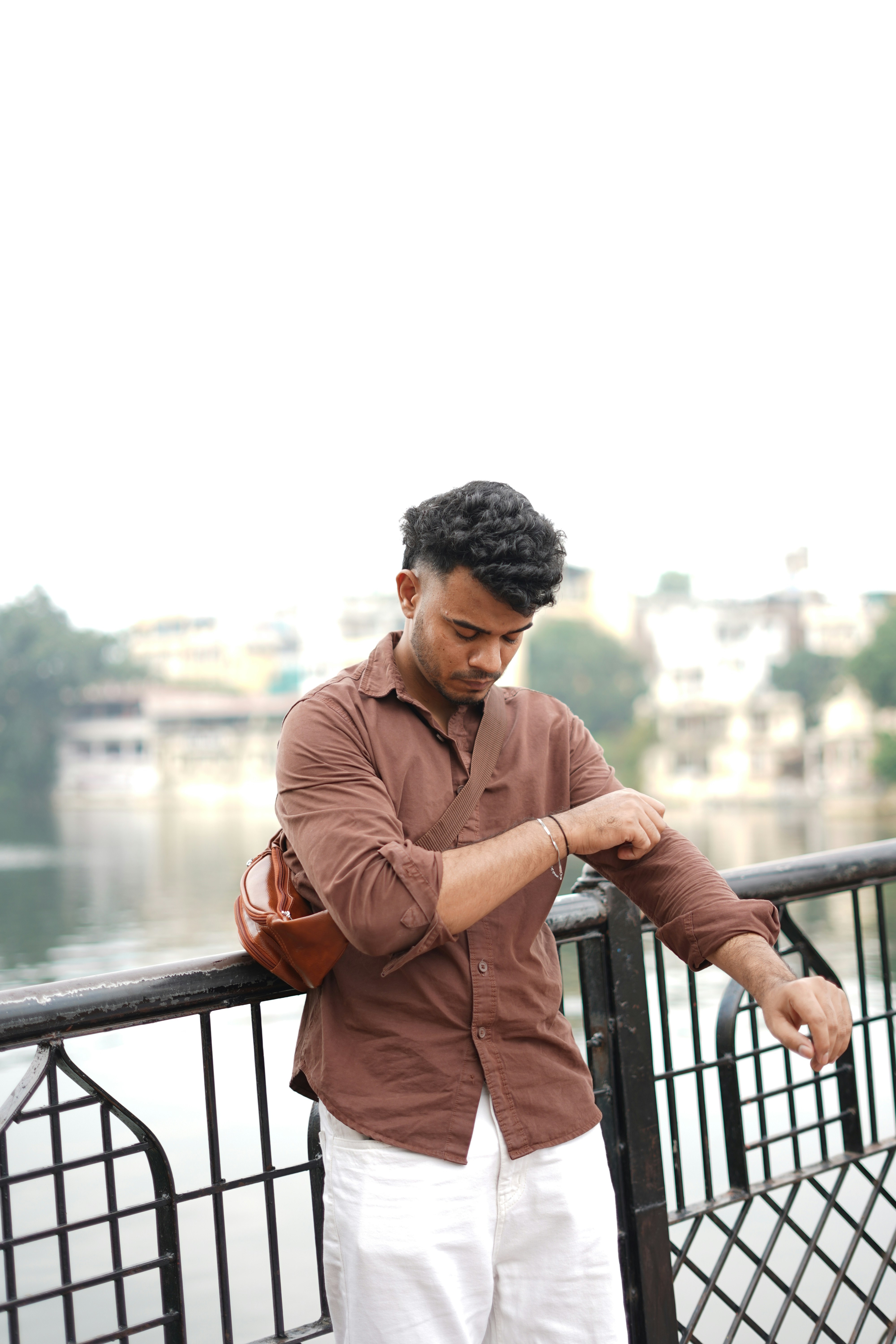 A young man in a brown shirt adjusts his sleeve while leaning against a railing, with a serene water backdrop and soft urban scenery.