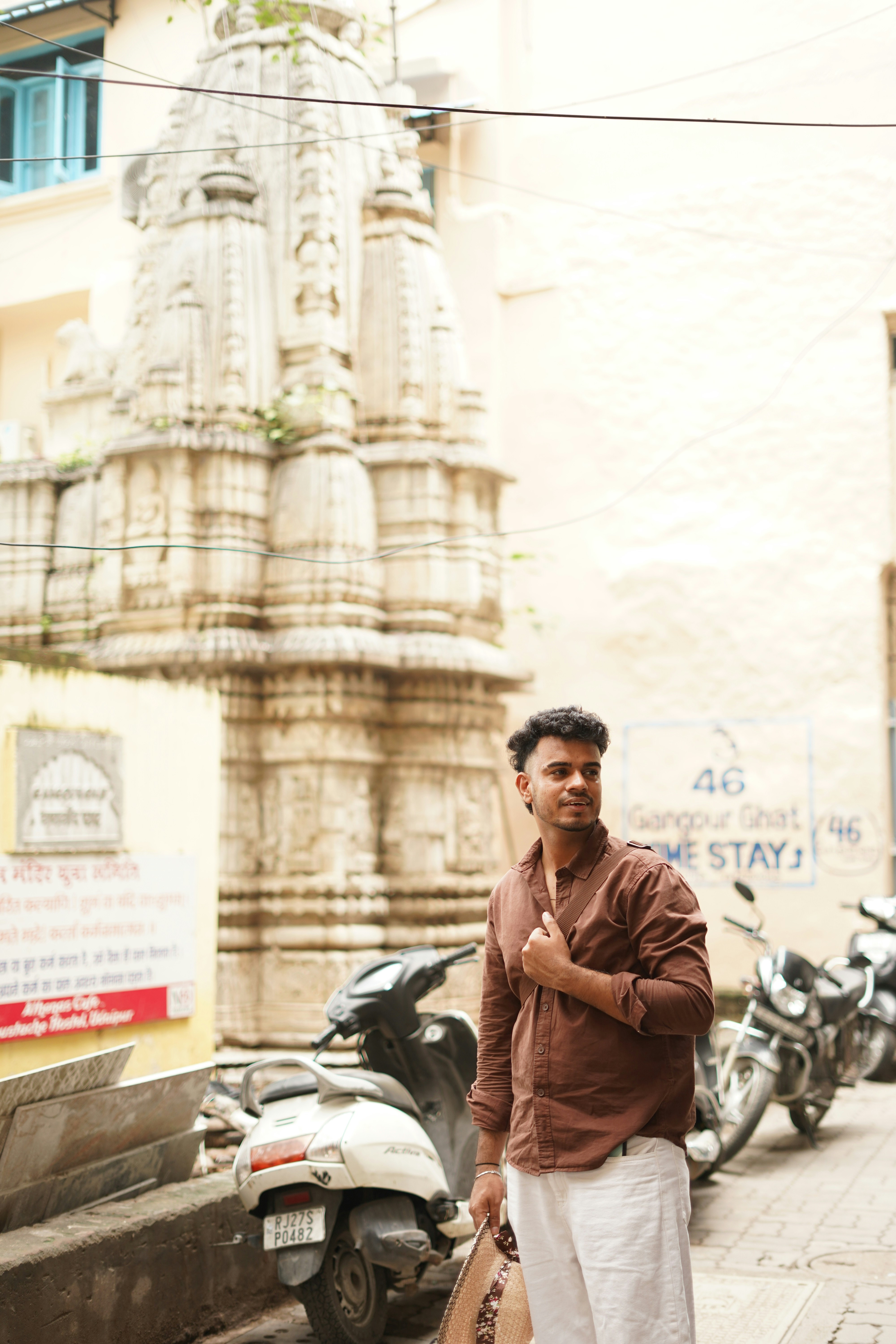 A man in traditional attire stands near an intricately carved temple, surrounded by parked scooters and vibrant street signs. The scene captures the blend of culture and modern life.